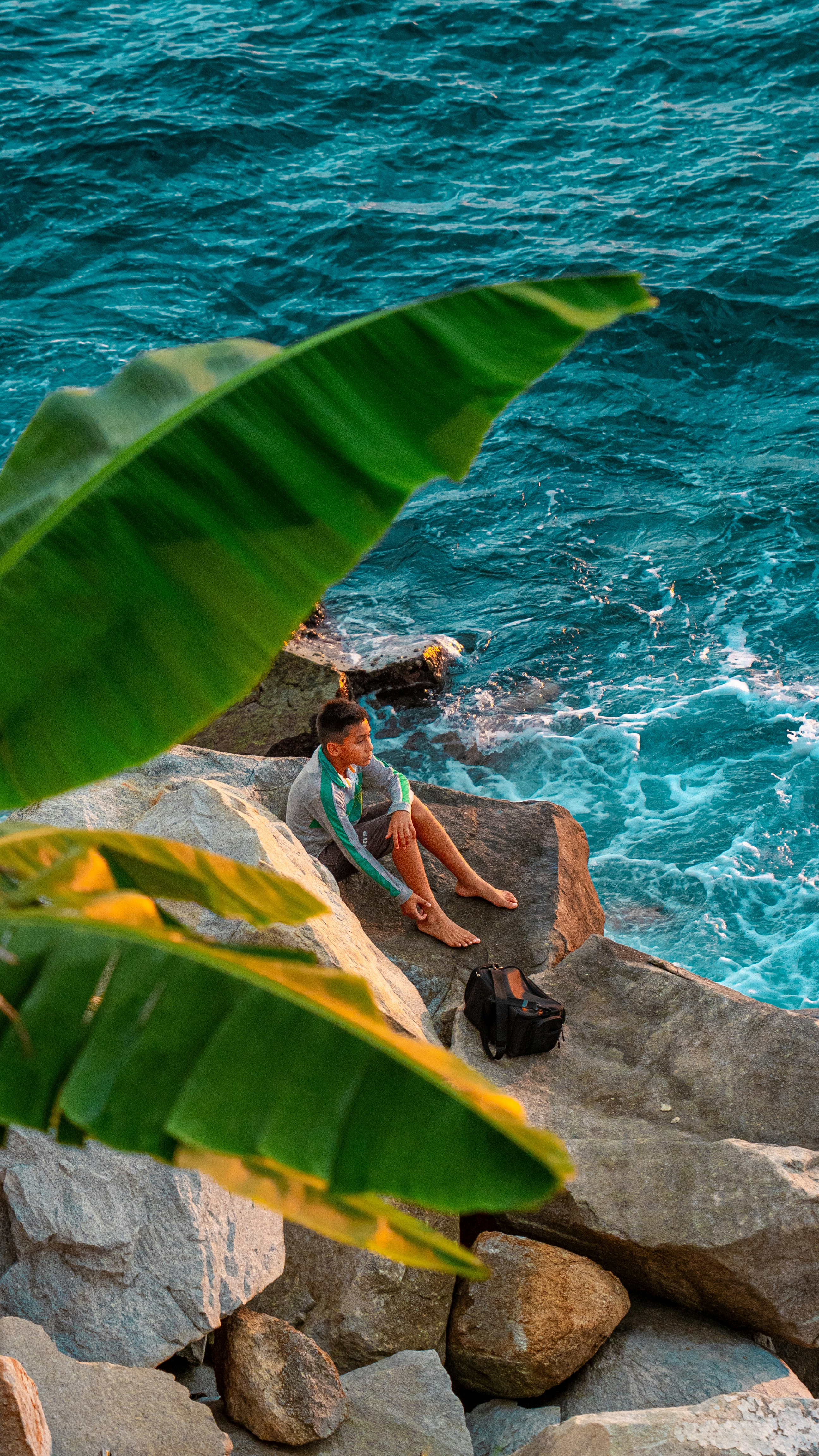 Man sits on rocky shoreline gazing toward the turquoise tide, a backpack resting nearby among large stones; tropical leaves frame the scene as waves crash below. (Photo is a coastal landscape with a human element.)