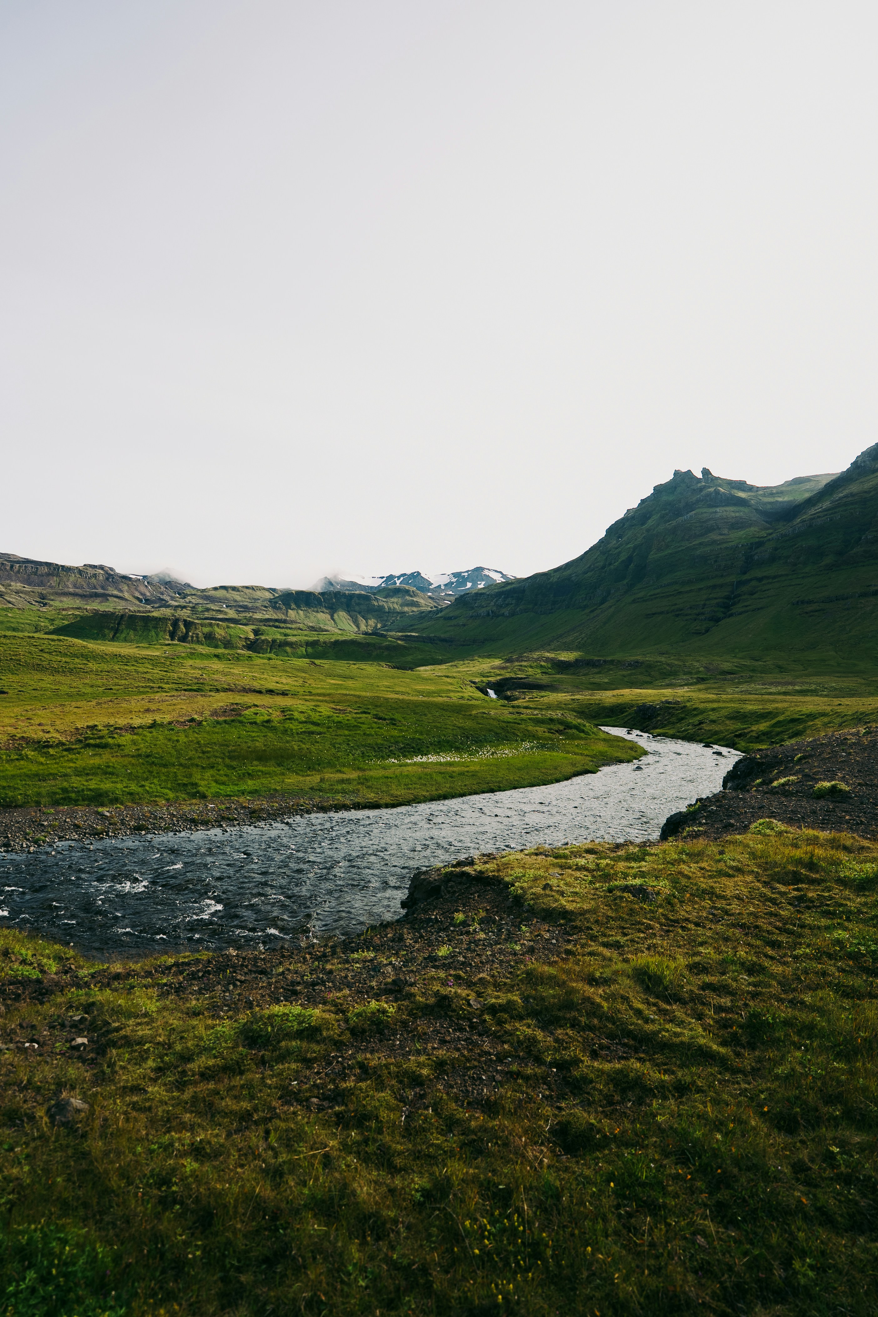 A winding river flows through a grassy alpine valley with distant hills under a pale sky; the scene emphasizes expansive landscape and gentle contours.