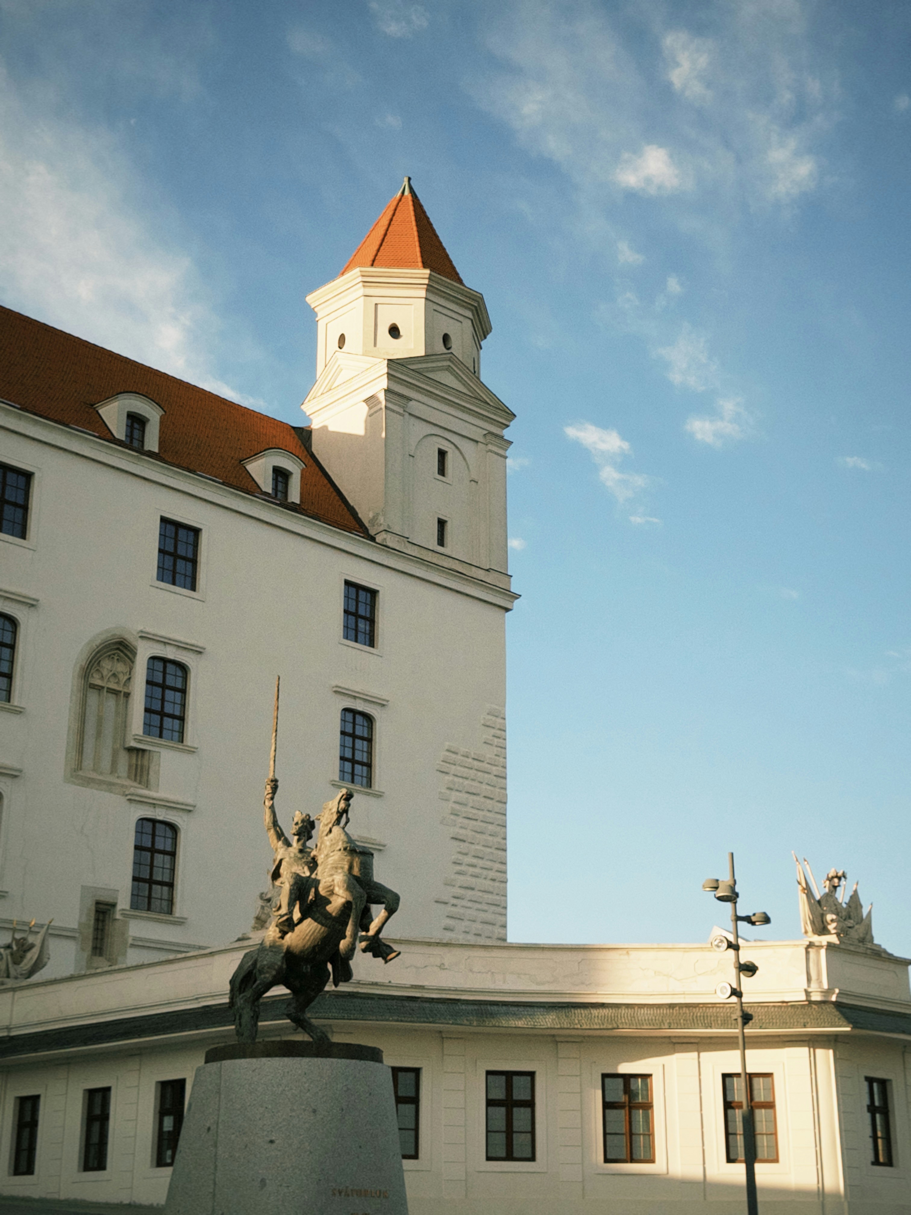 Sunlit castle wing with statue of a rider on horseback in the foreground against a clear blue sky. The scene emphasizes classic architecture and sculptural detail.