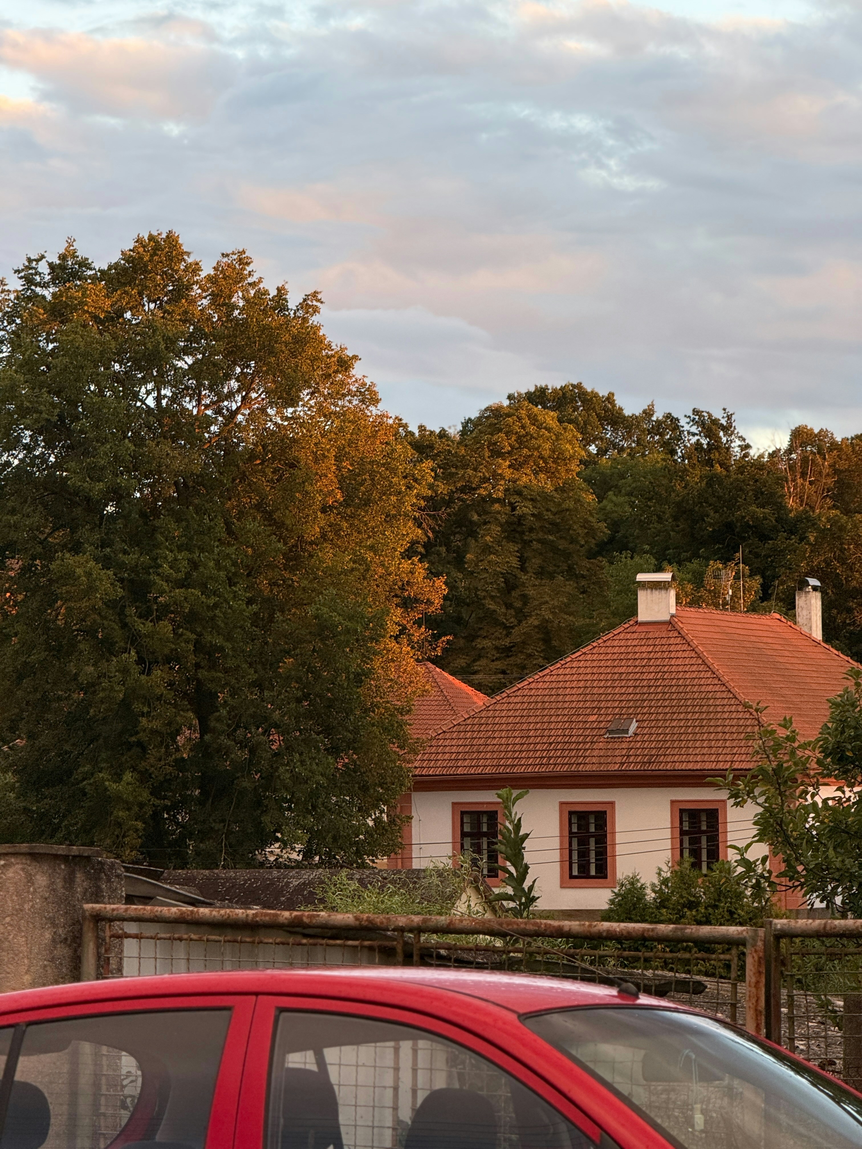 Red car at the edge of a sunlit courtyard with a brick house and autumn trees