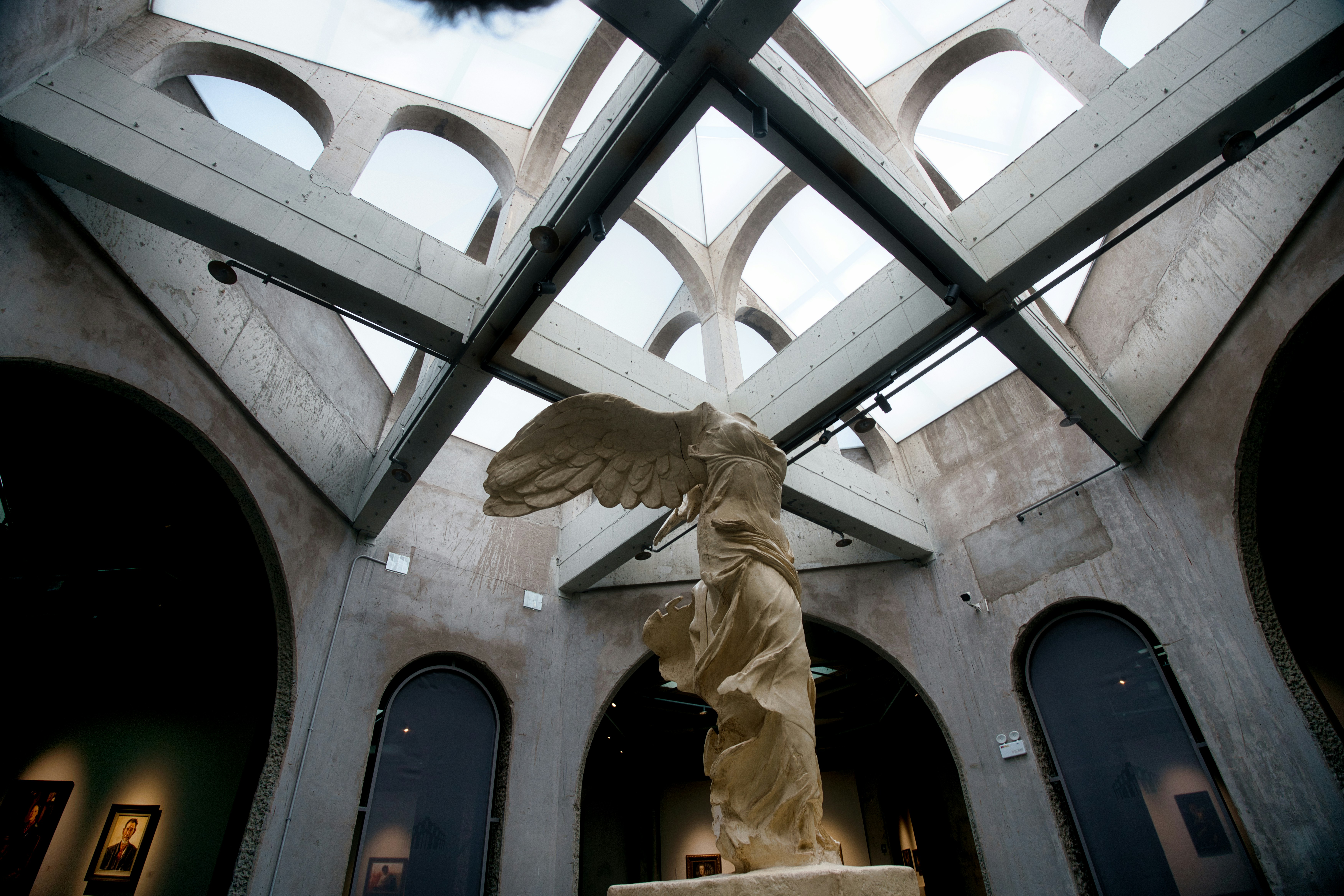 Winged statue illuminated within a museum atrium, framed by arched windows and a geometric skylight.