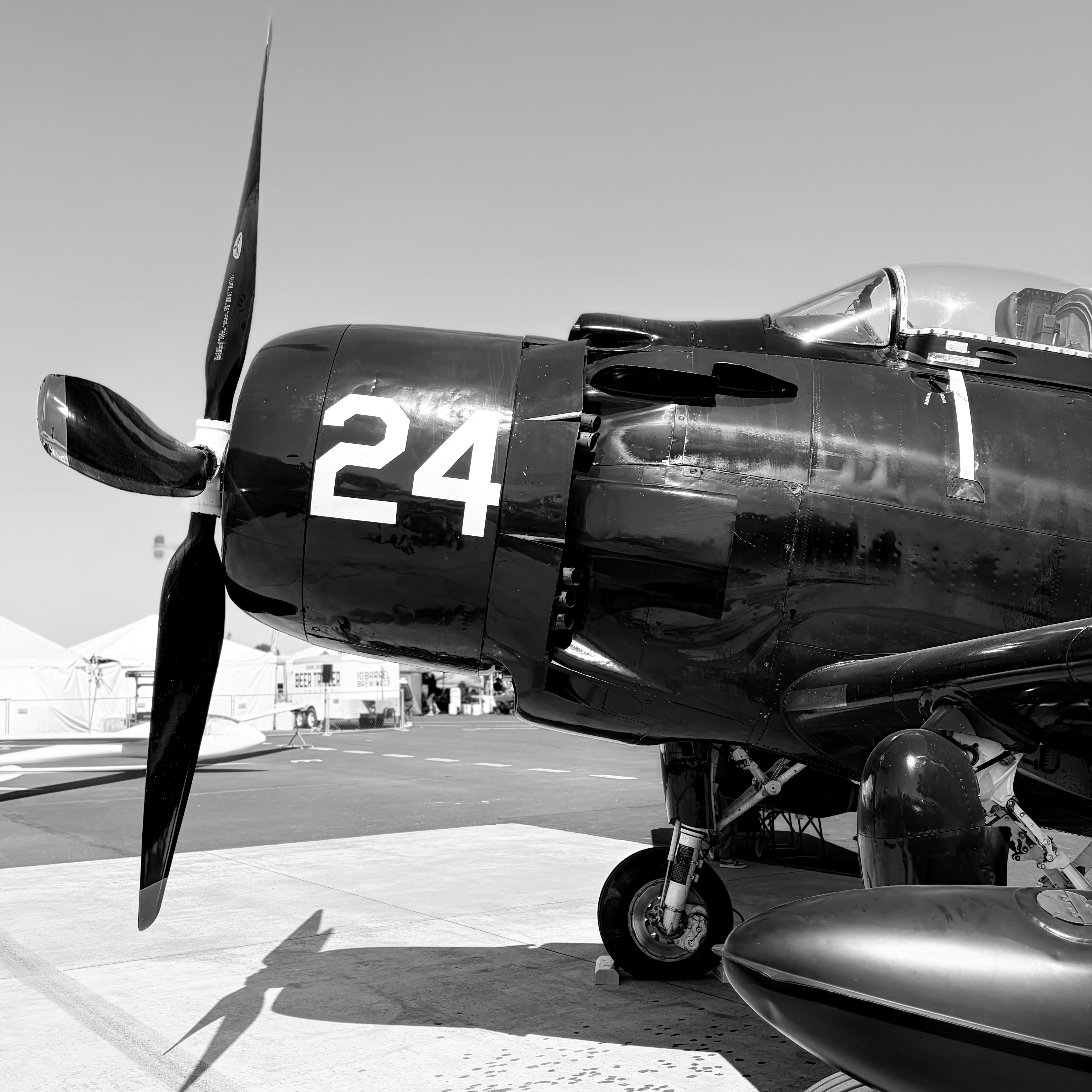 Close-up of a black vintage aircraft nose and propeller with the number 24, parked on a tarmac; high-contrast grayscale emphasizing metal texture and weathered surface.