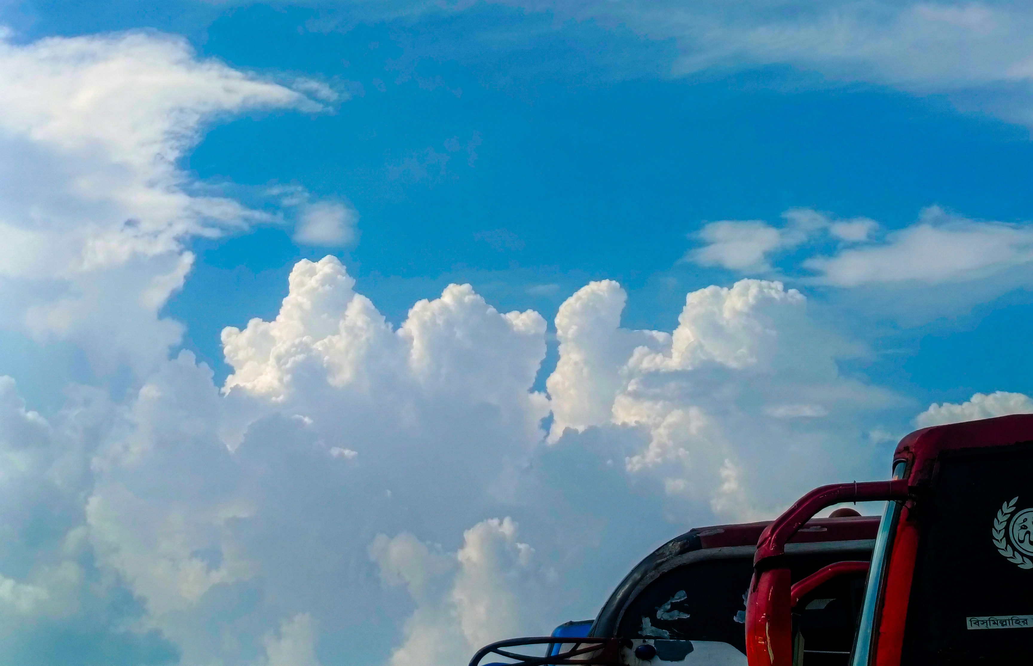 Bright blue sky with towering cumulus clouds above a red rooftop cluster; minimal foreground elements foreground the airy clouds and sunlit shapes.