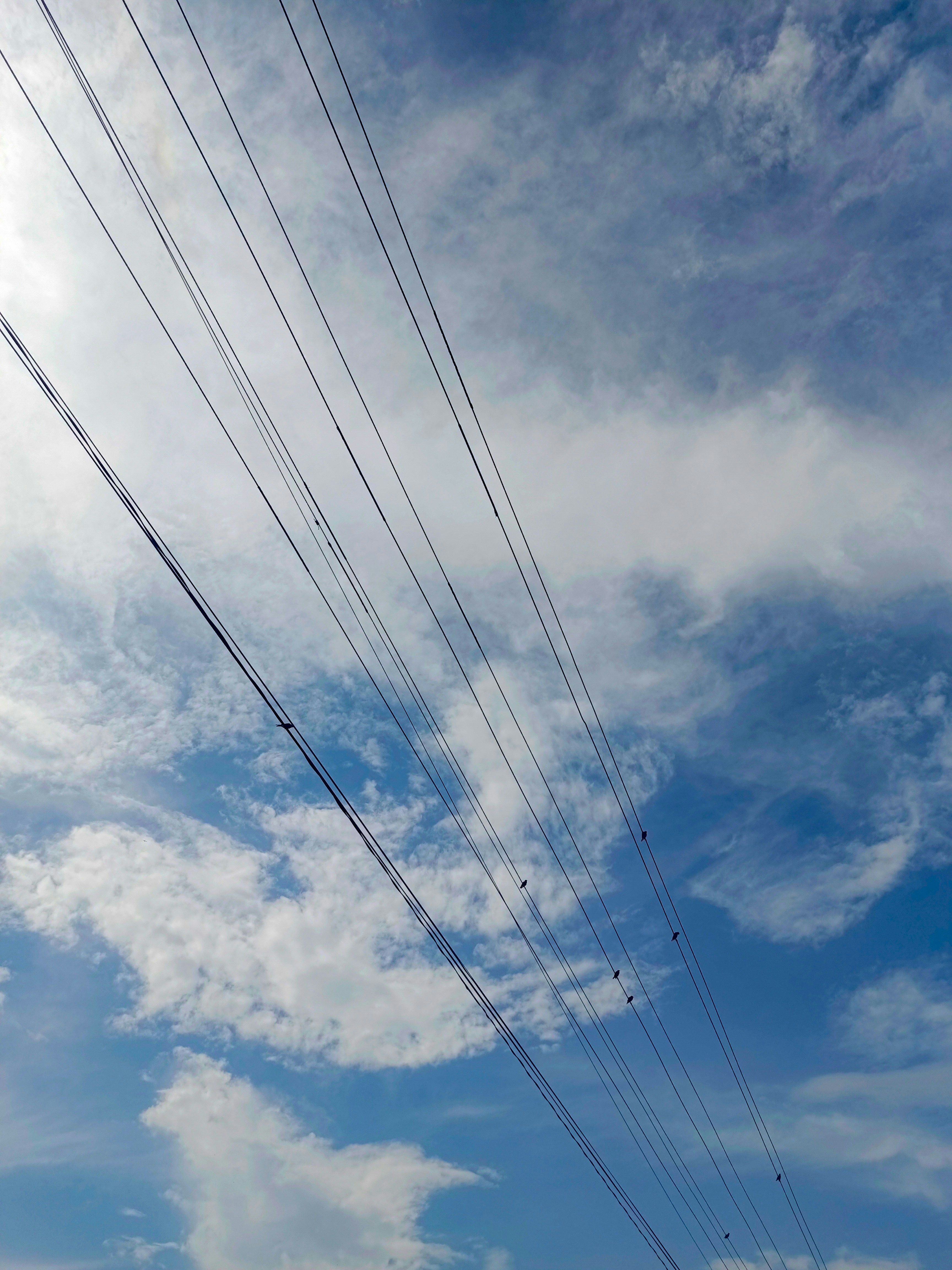 Bright sky with diagonal power lines slicing across fluffy clouds, creating a geometric contrast against the blue backdrop. Minimal foreground details, emphasizing depth and lines in the composition.