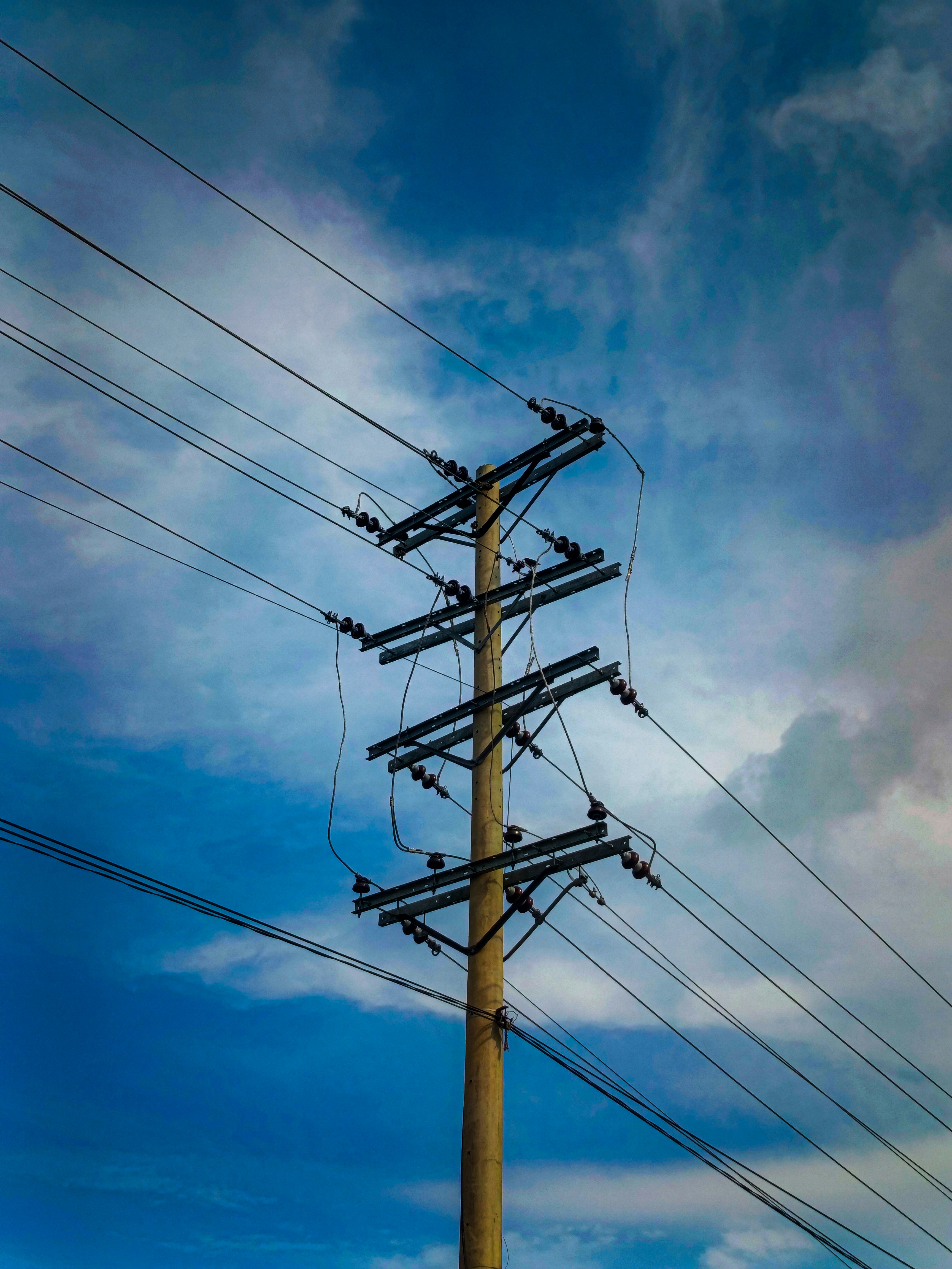 A tall utility pole with multiple crossarms and wires stretches upward against a blue sky with scattered clouds, emphasizing urban infrastructure. Photograph highlights structural geometry and contrast between man-made lines and natural atmosphere.