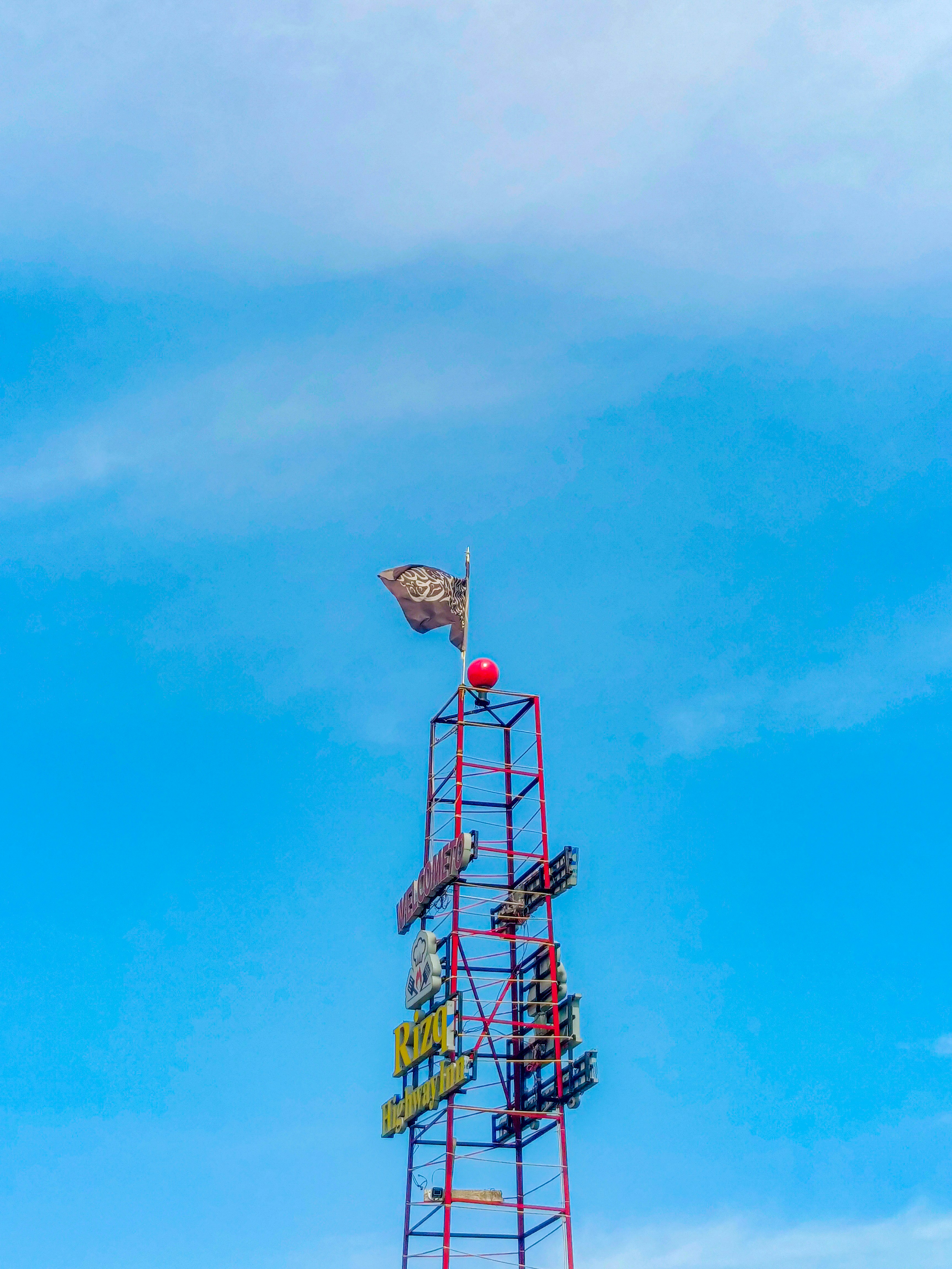 Towering red framework with a flag and a red orb atop, set against a bright blue sky; signs hang below, suggesting a roadside landmark or amusement-park structure.