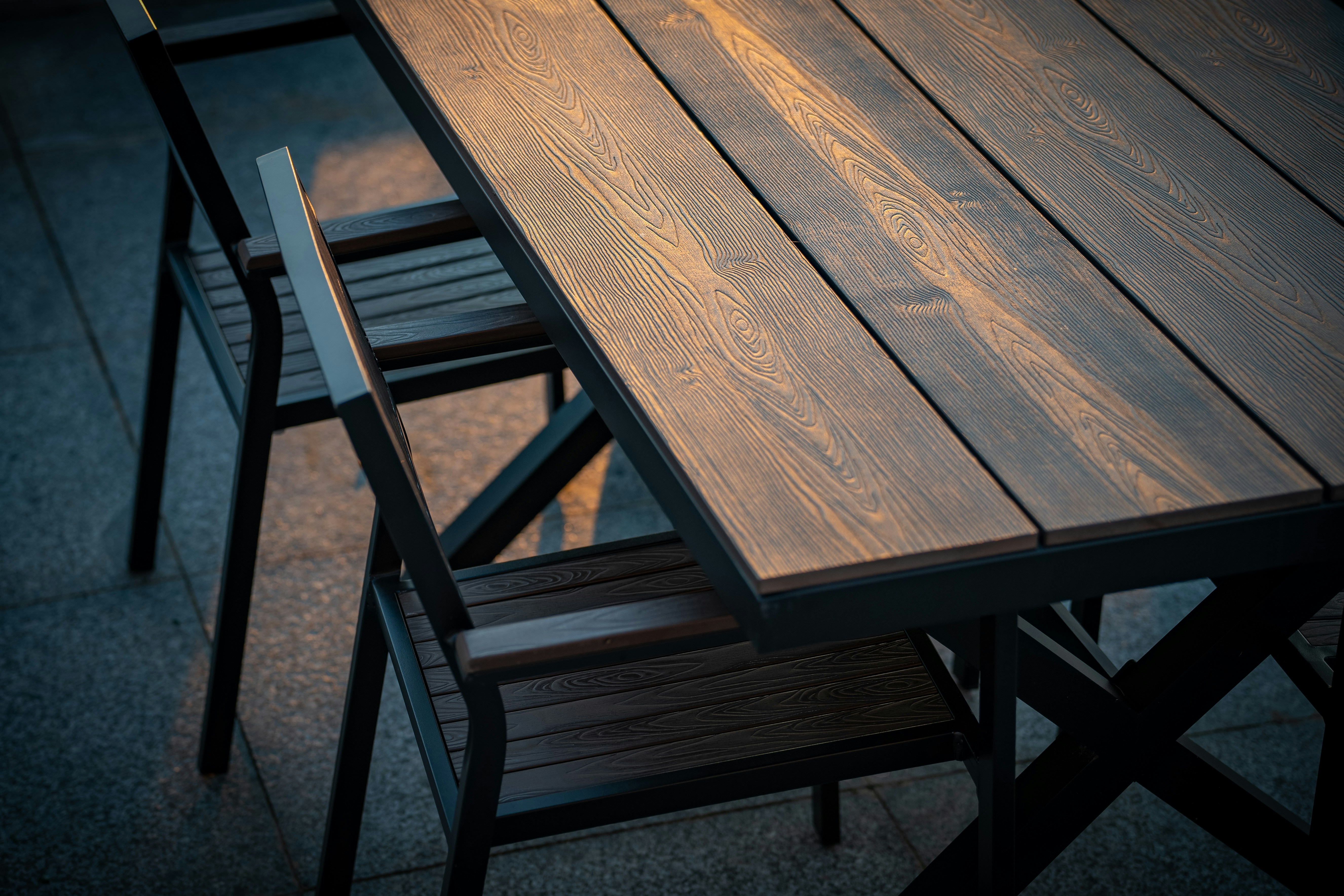 Wooden outdoor dining table with slatted chairs, captured in soft, warm light showing rich grain and subtle shadows.