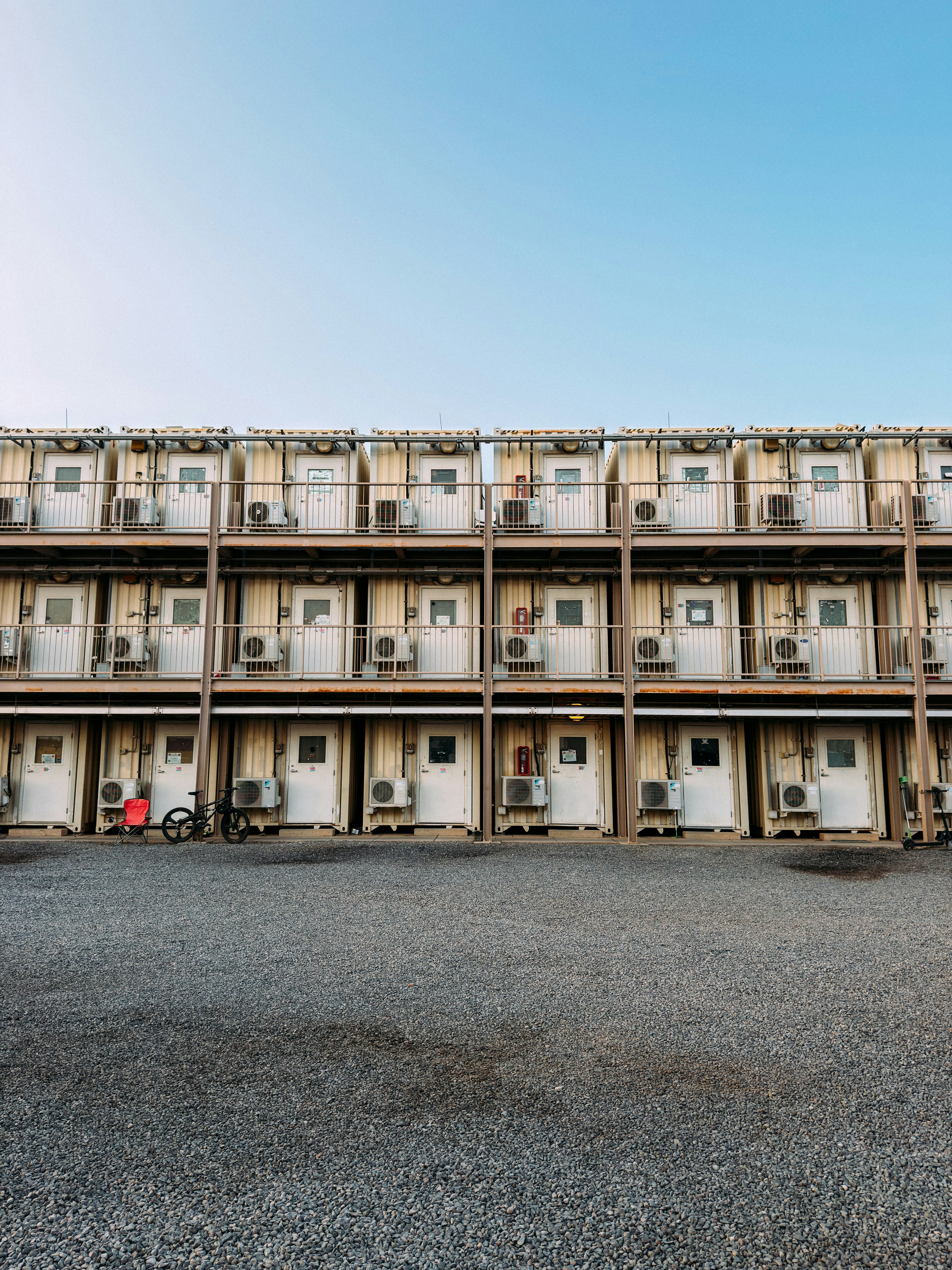 Row of motel units with balconies and air conditioning units, set against a pale blue sky; ground level shows gravel and a lone bicycle. A clipped red chair hints at lived-in scenes.