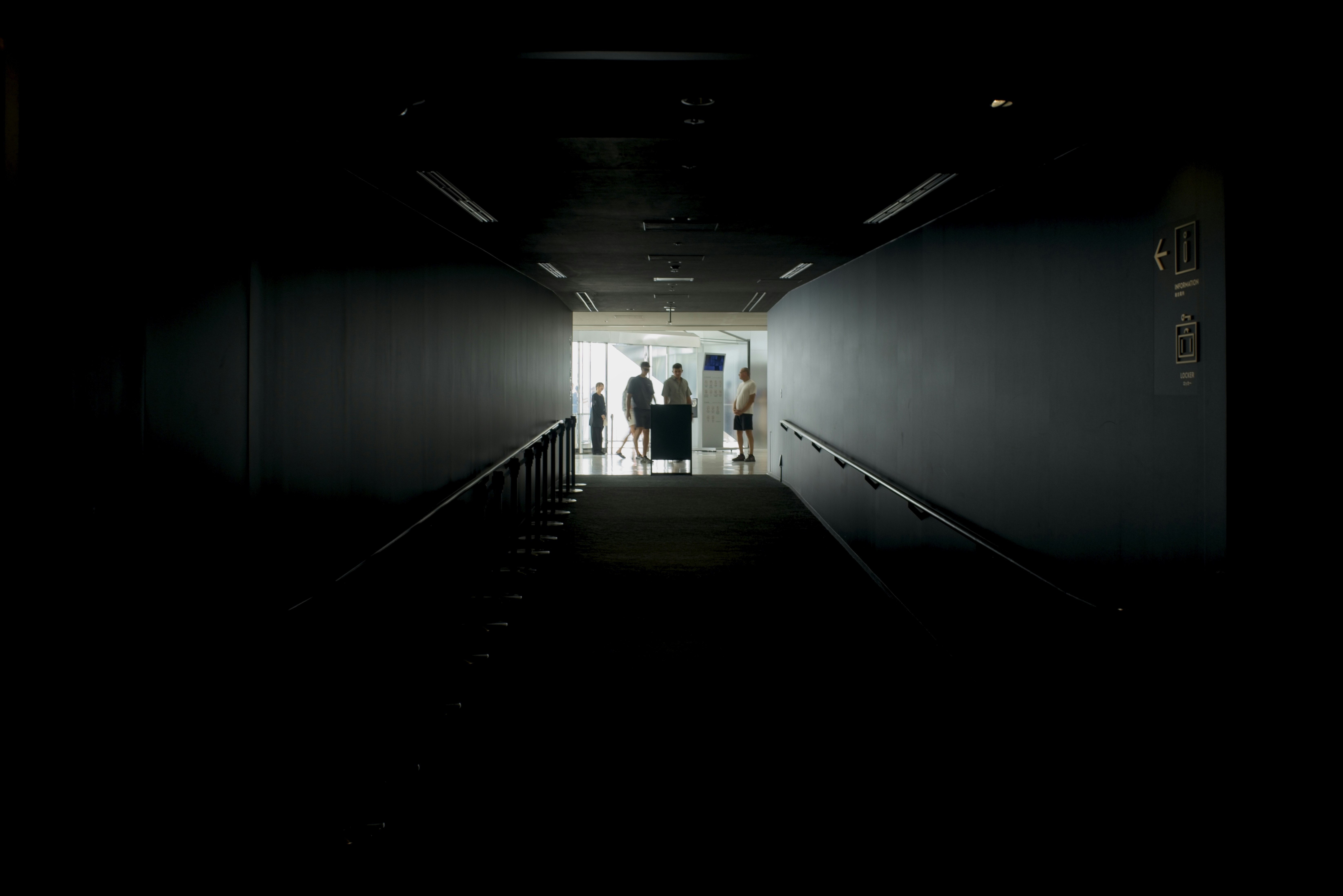 A quiet, moody shot down a long, dark corridor leading toward Shibuya Sky's bright entrance hall. Handrails line either side, guiding the eye toward a distant doorway where a few silhouetted figures pause near a locker area and information point. Soft daylight spills in from the glass entrance beyond, contrasting sharply with the deep shadows of the tunnel-like hallway. The composition's strong symmetry and dramatic light-to-dark gradient create a sense of quiet anticipation, capturing the transitional moment just before visitors emerge onto Tokyo's rooftop skyline view.
