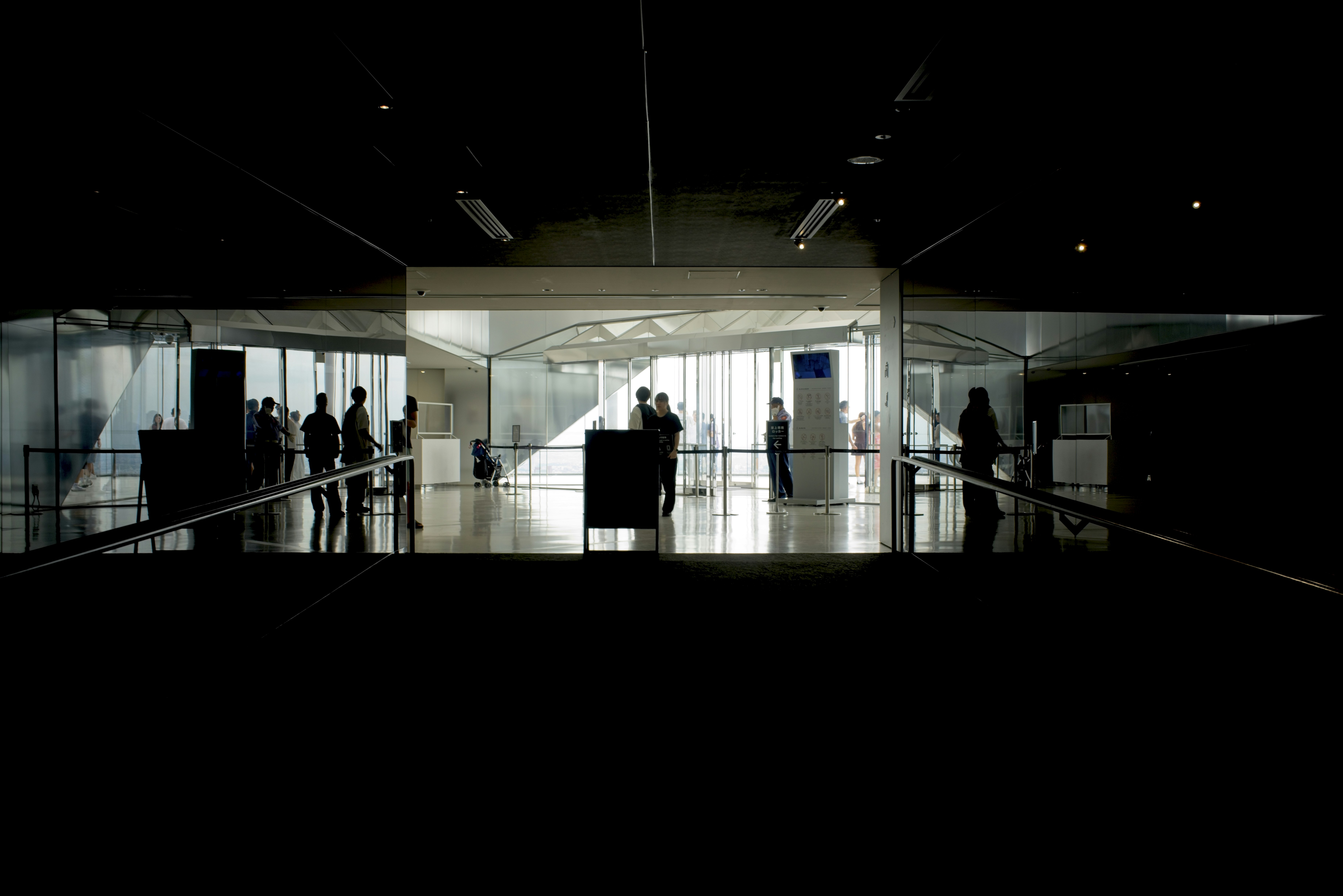 From a shadowed entrance hallway, this frame opens onto Shibuya Sky's bright glass lobby, its mirrored walls creating a symmetrical, almost kaleidoscopic effect. Silhouetted visitors move through the sunlit interior, past a stroller and directional signage guiding guests toward the rooftop deck. Diffused daylight floods in through floor-to-ceiling windows, softening the architectural lines beyond. The strong contrast between the dark foreground and glowing background lends the composition a graphic, cinematic feel, quietly capturing the steady rhythm of visitors passing through on a hazy Tokyo