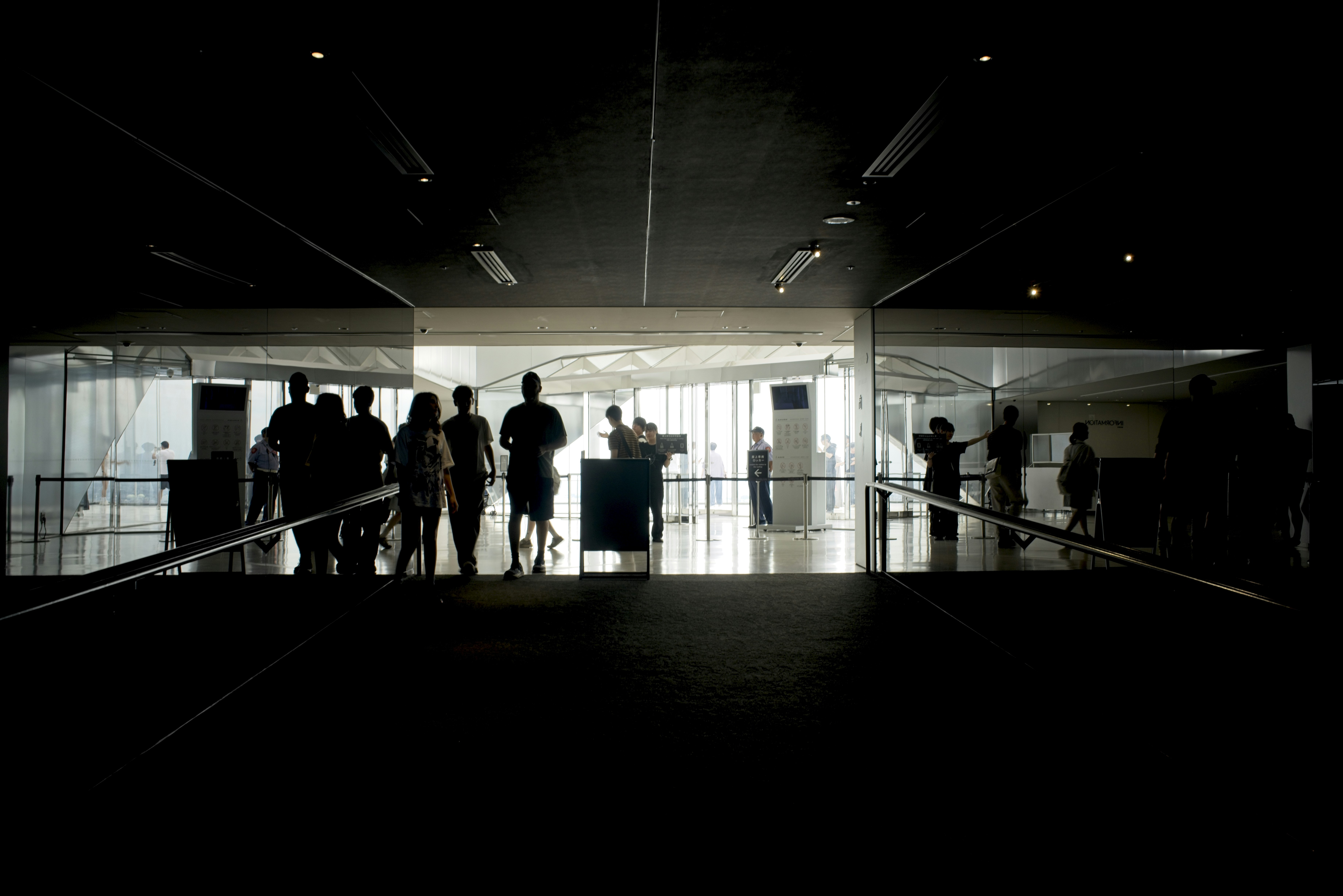 A cluster of visitors walks in silhouette toward Shibuya Sky's radiant glass lobby, framed by a dark, mirrored hallway that doubles the scene symmetrically on both sides. Backlit by strong daylight, the group's outlines stand out sharply against the bright interior, where more travelers, a security guard, and information signage populate the space. The bold contrast between shadow and light gives the image a graphic, almost cinematic energy — a candid glimpse of the steady flow of visitors converging toward Tokyo's rooftop observation deck on a hazy July afternoon.