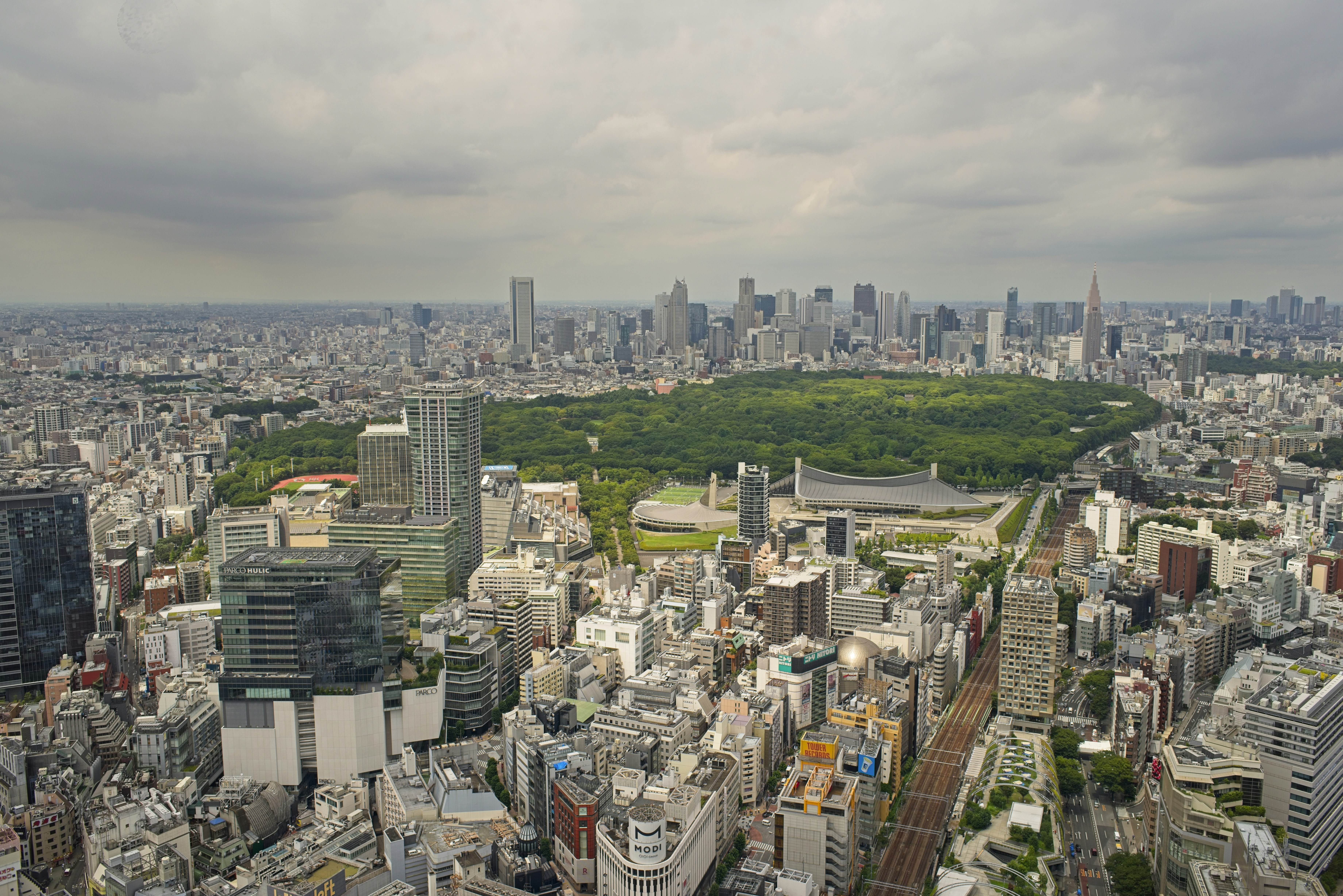 Looking out from Shibuya Sky toward Shinjuku, this view captures Tokyo's vast urban sprawl framed by the deep green expanse of Yoyogi Park below. The stadium's curved roofline stands out amid the trees, while straight railway lines slice through the surrounding neighborhoods. A grey, humid July sky hangs low over the distant skyscrapers, softening the skyline into a hazy silhouette. Closer buildings reveal Shibuya's dense commercial rooftops in sharp detail, creating a layered composition that showcases both the city's relentless density and its pockets of preserved nature.