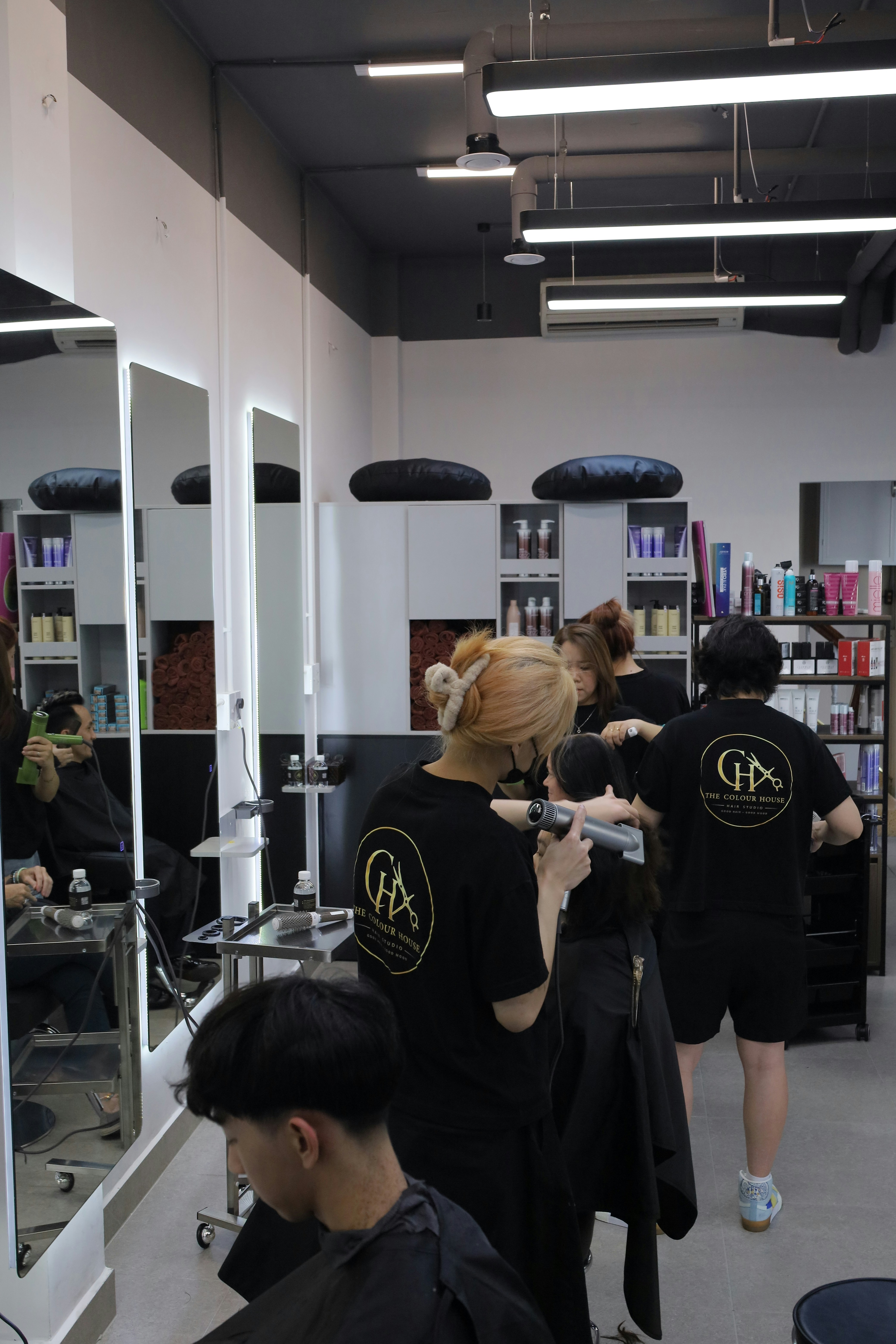 Hair stylists work on clients in a busy salon, mirrors reflecting activity and shelves stocked with products in a modern space. This image captures a candid moment of styling and attention to detail in a professional setting.