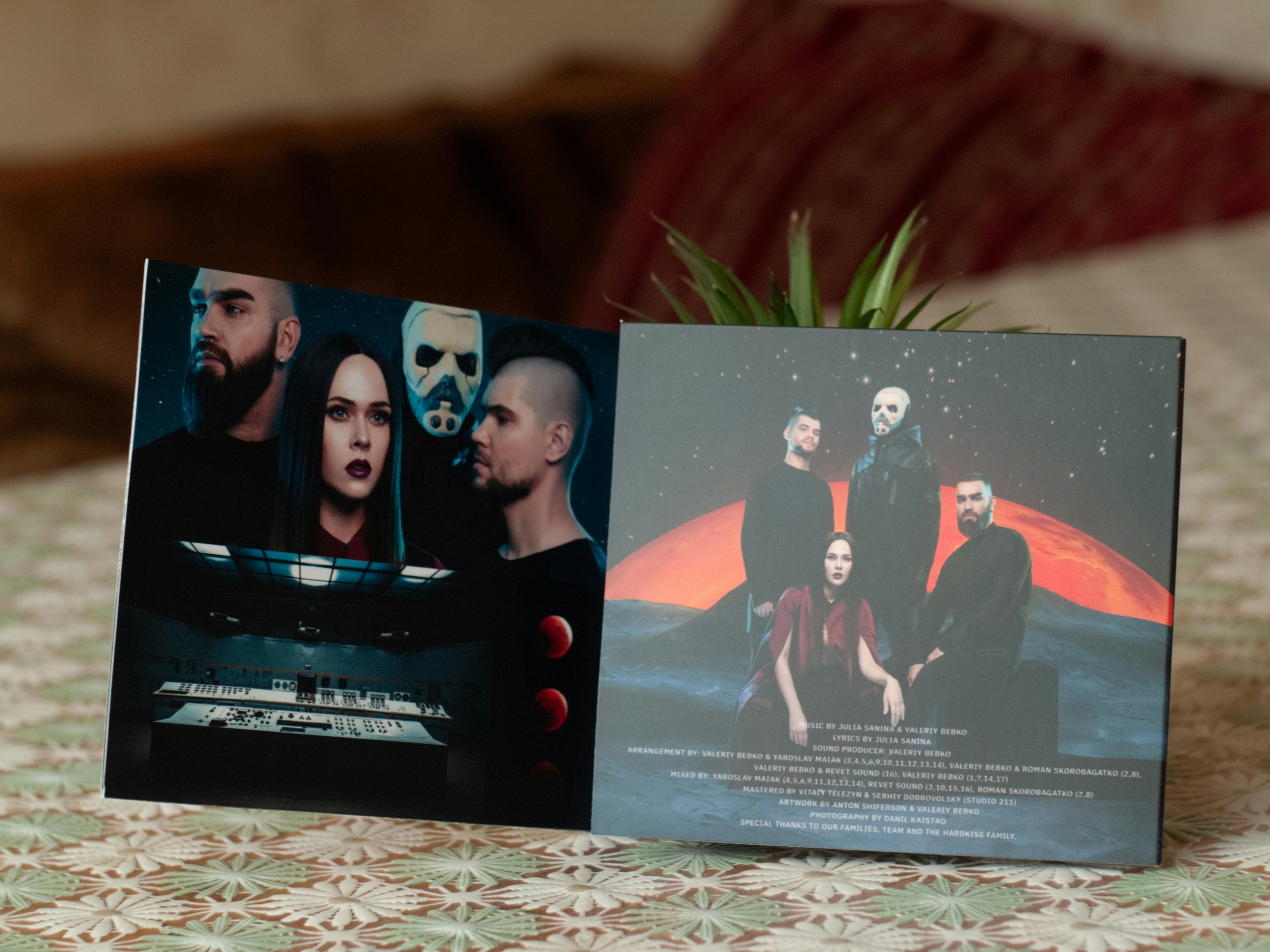 Two vinyl-style sleeves rest on a patterned surface, the left panel showing a trio of faces and the right panel featuring a multi-person group against a lunar backdrop.