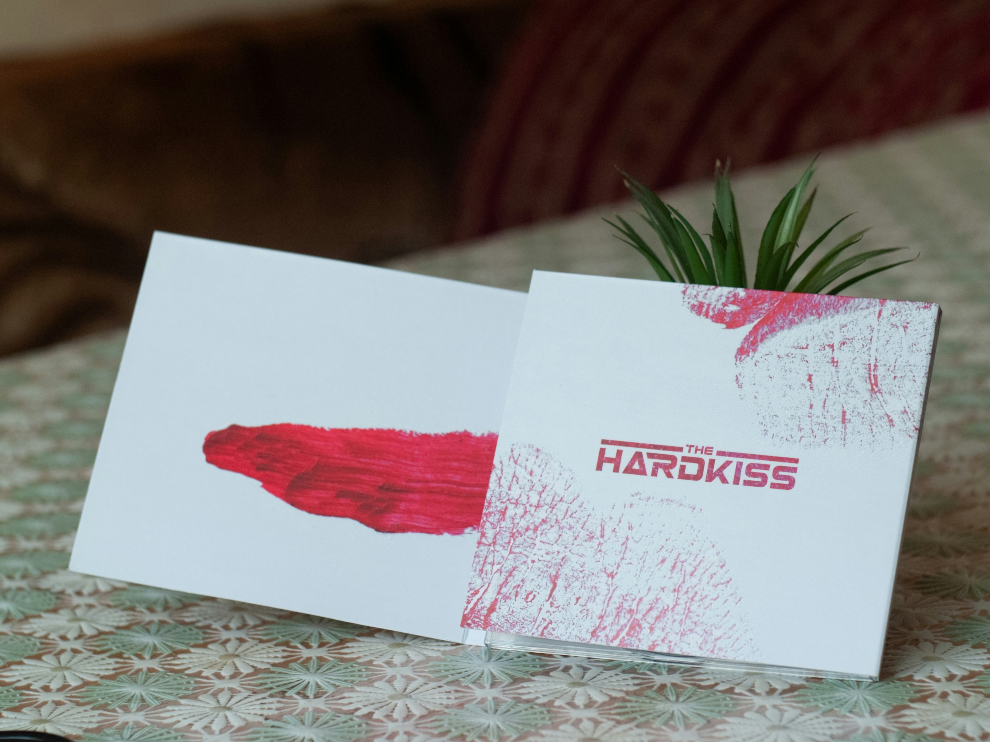 Two white cards with a red brushstroke design rest on a patterned surface; a small green plant peeks from behind, suggesting a staged still-life or product presentation.