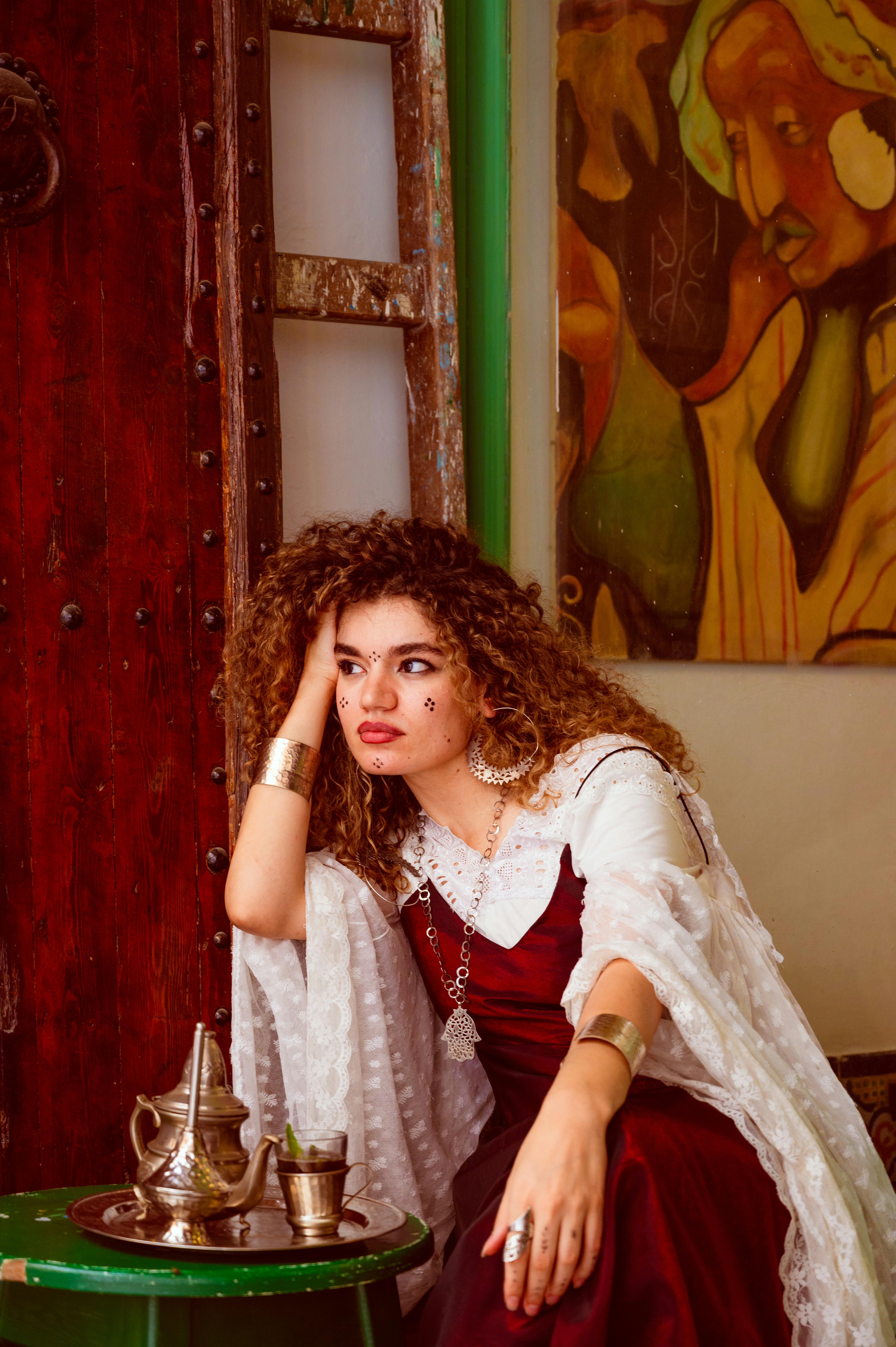 A woman in a red dress draped with lace sits beside a green table with silver tea set, leaning her head on her hand and gazing off-frame; painterly mural and rich wood backdrop provide warm, vintage atmosphere.