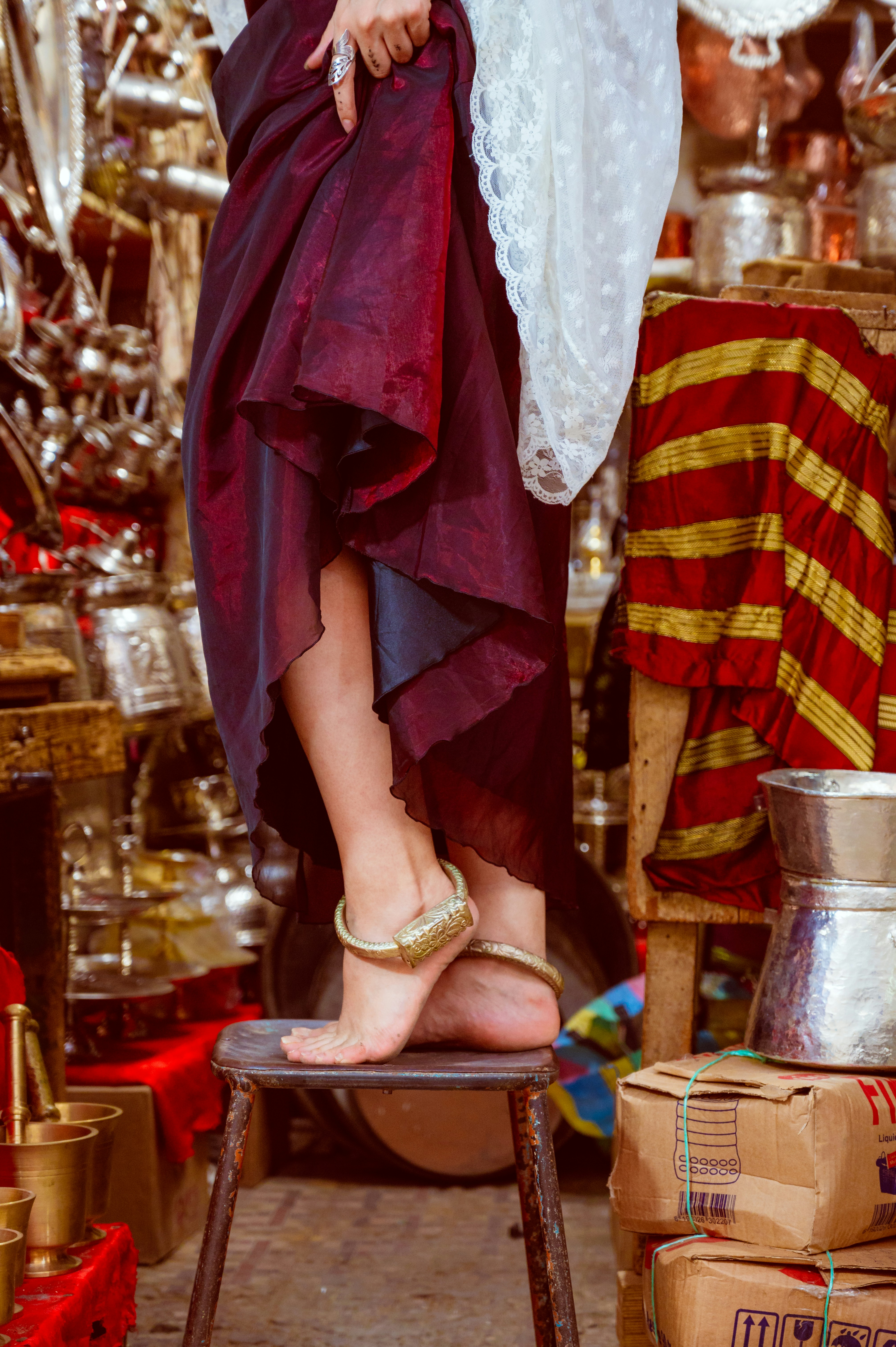 Bare feet and ornate sandals hover on a rusted stool amid a bustling market, with layered garments and metallics creating a warm, textured scene.