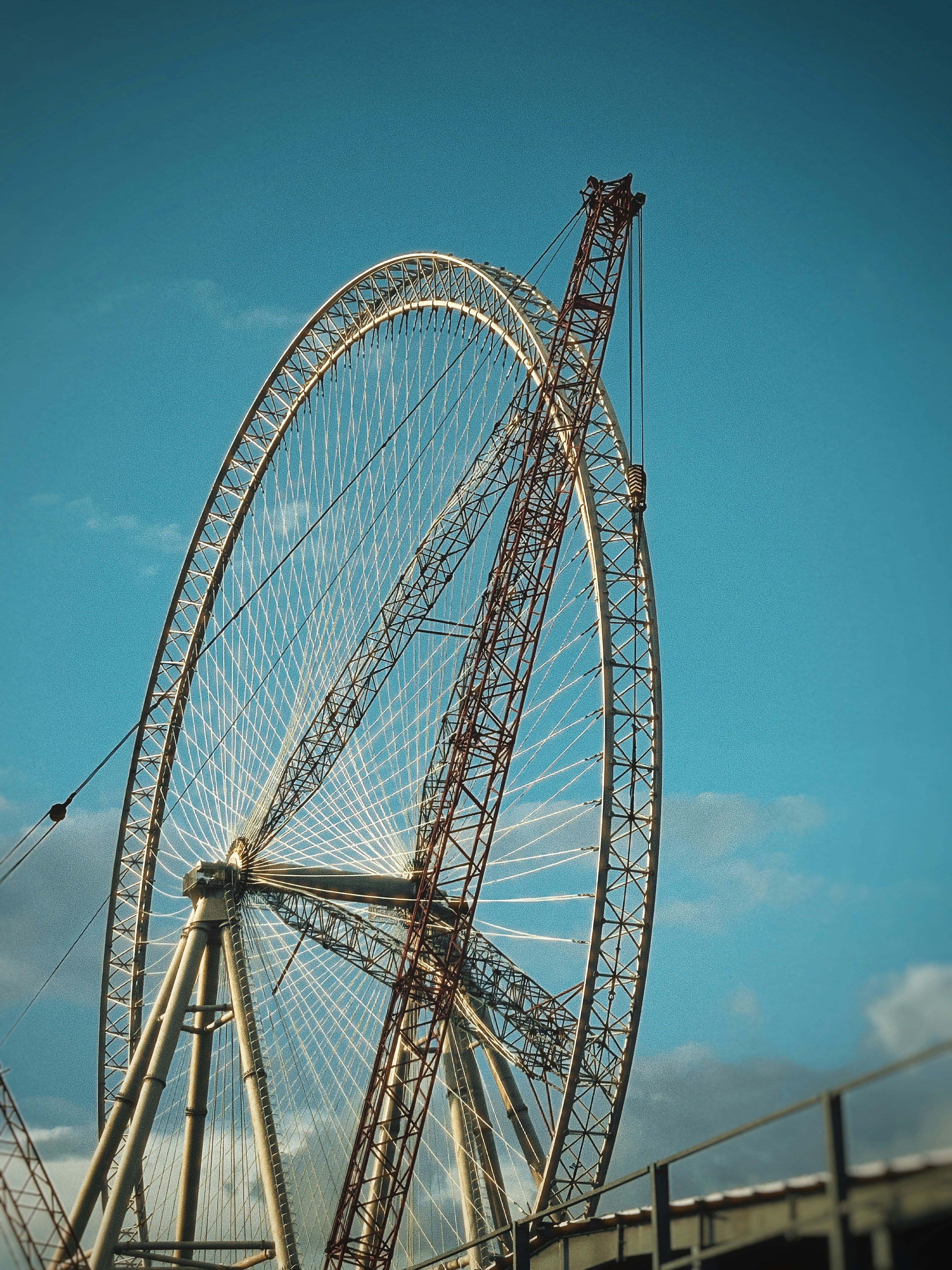 Ferris wheel under a clear blue sky, with structural supports in the foreground and subtle clouds in the background.