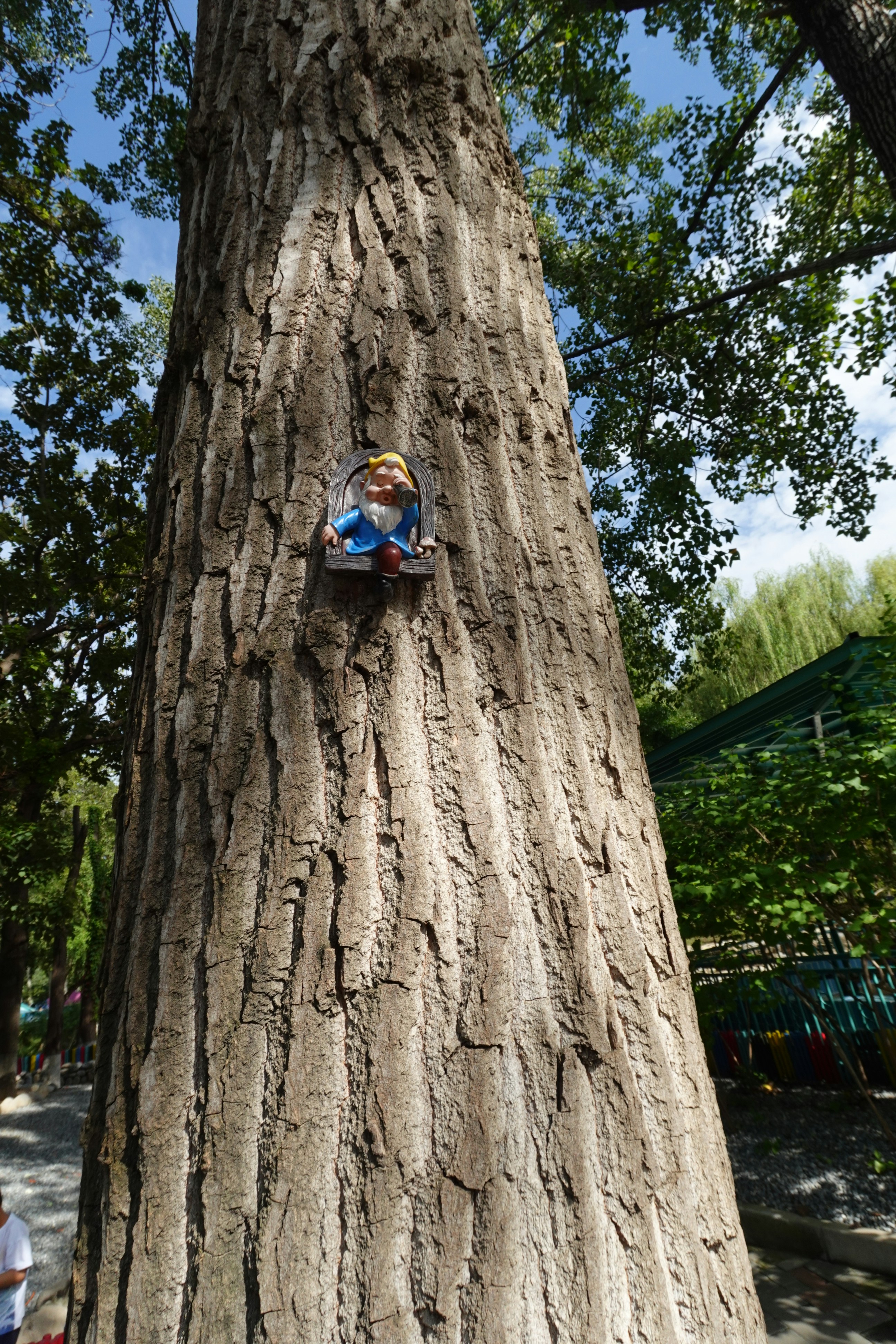 A small figurine on a swing is attached to the trunk of a towering tree, set against a sunny park scene with greenery and a distant shelter visible.