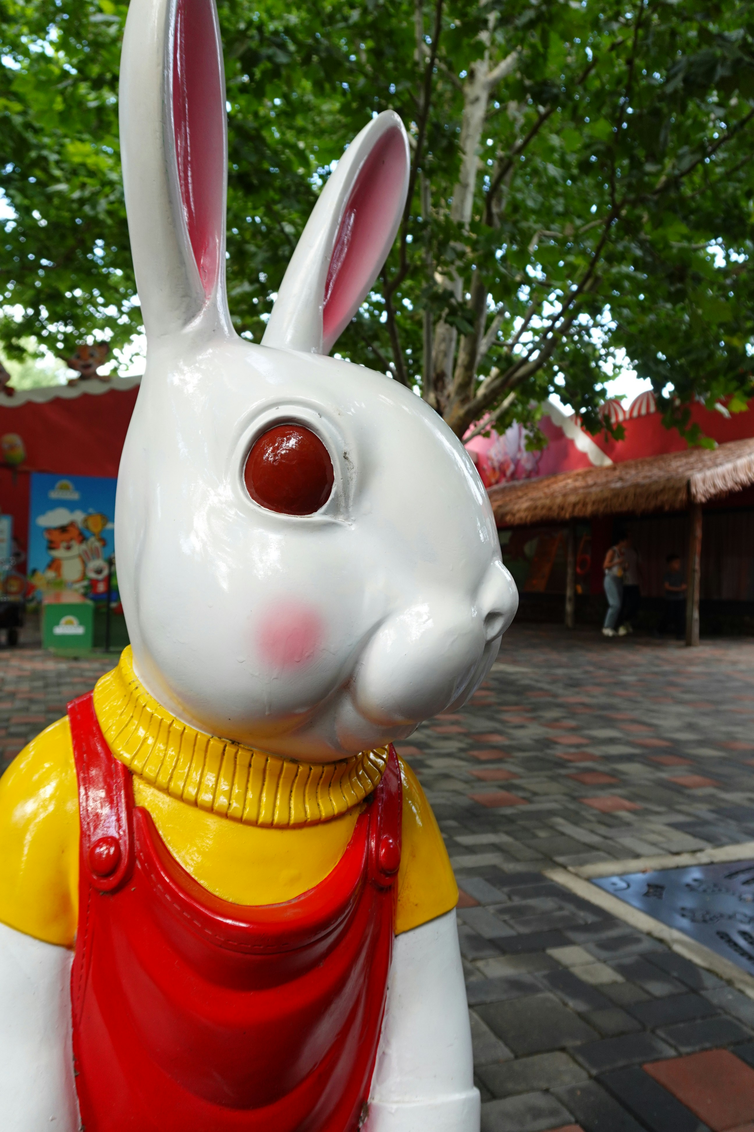 Bright white bunny statue with red eyes and rosy cheeks, wearing a yellow shirt and red overalls in an outdoor park setting.