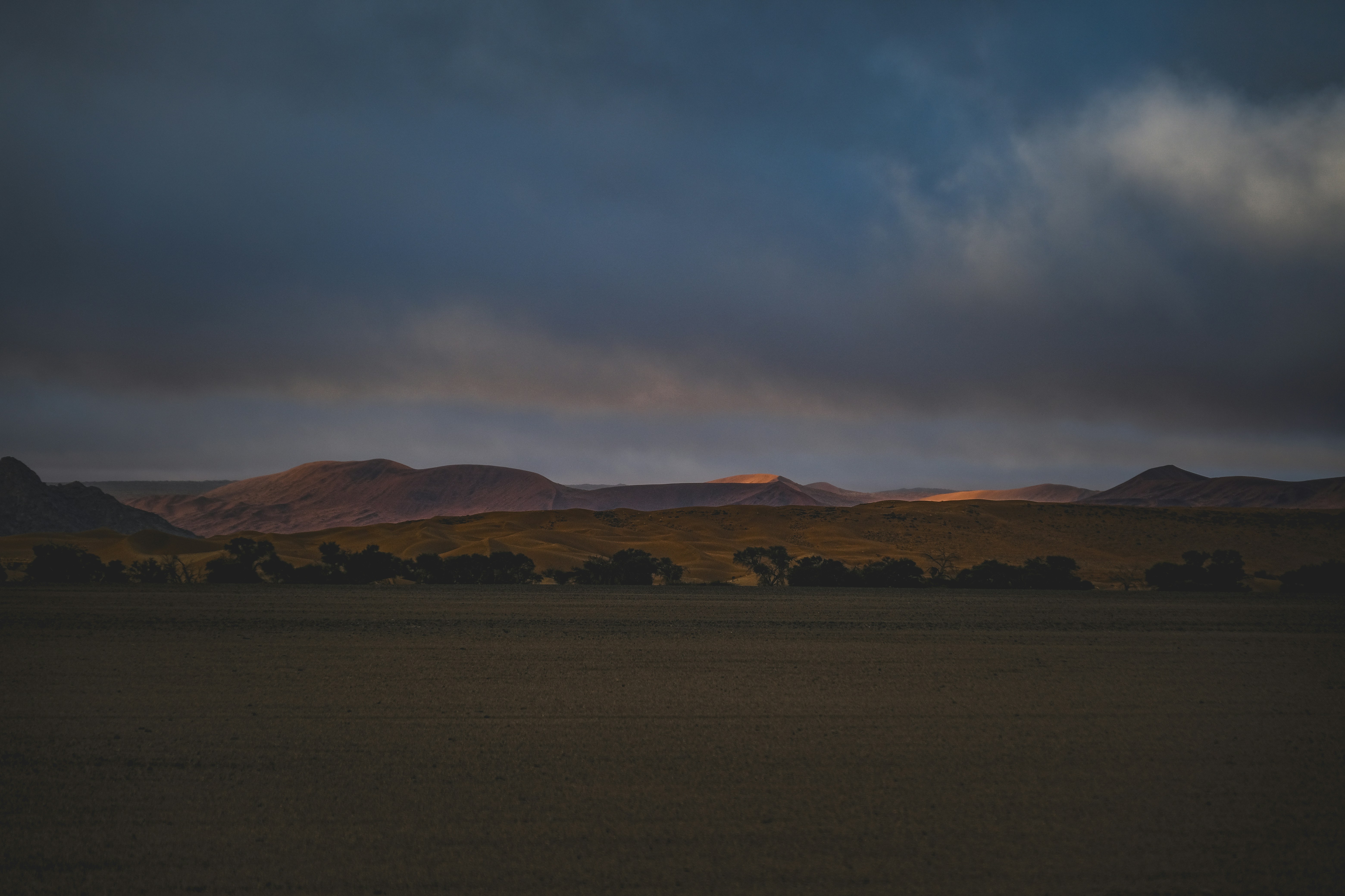 Low-contrast landscape with distant rolling hills under a moody sky; a narrow band of warmth highlights the ridge, while the foreground remains dark and expansive.