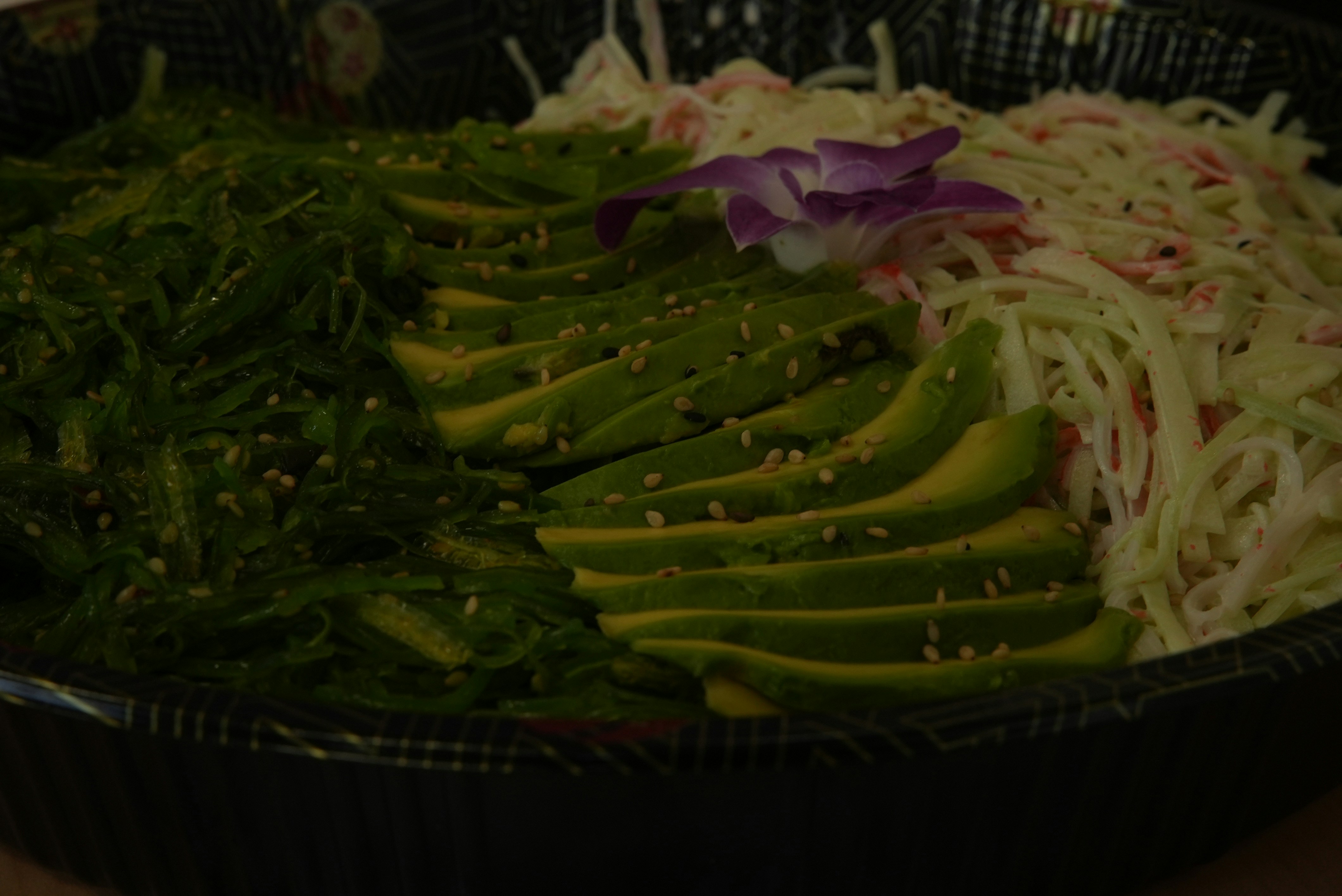 A close, dimly lit platter of sliced green peppers and white onions arranged in a circular bed with sesame seeds and an orchid garnish; a culinary presentation focusing on texture and color variety.