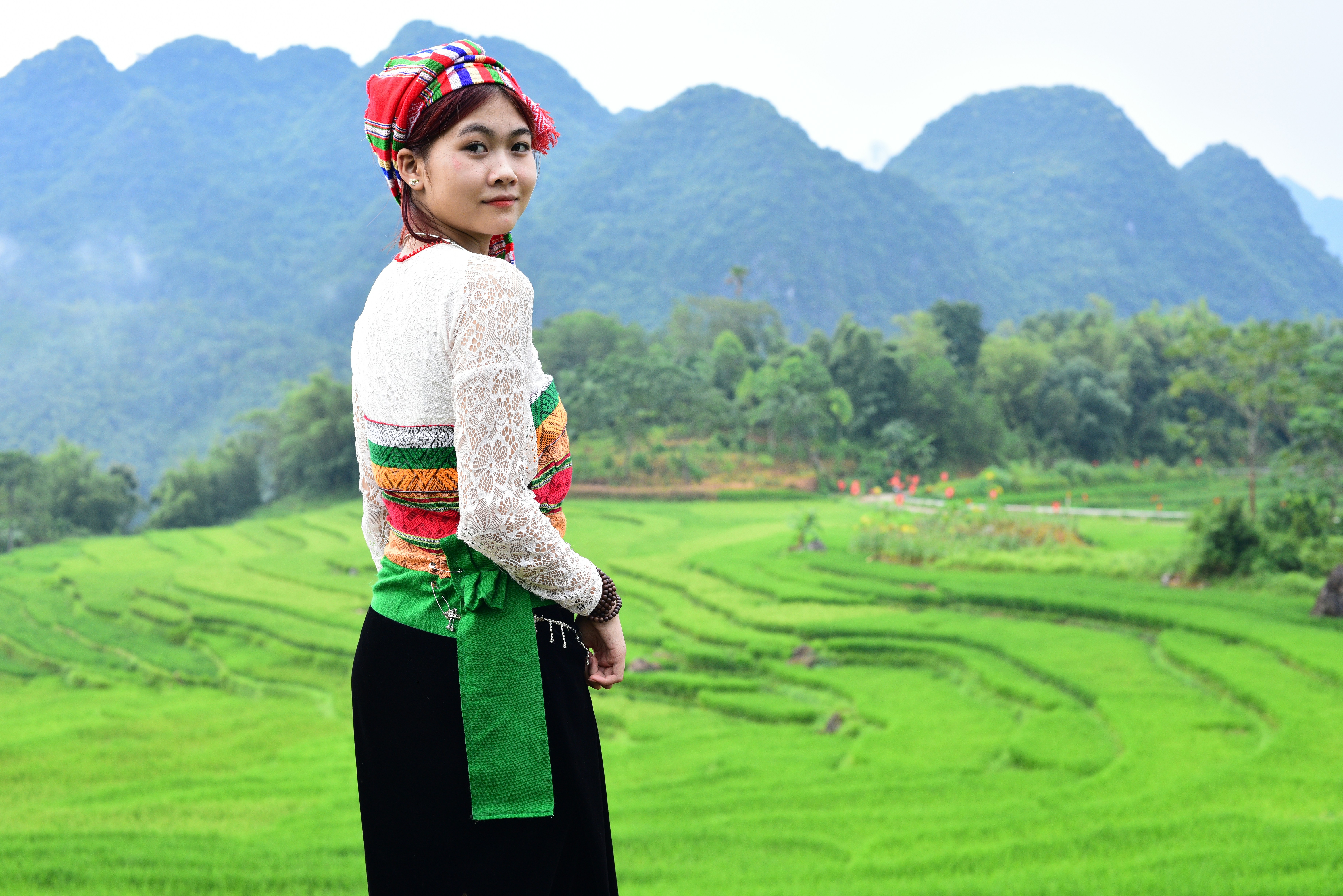 A woman in traditional attire stands in front of emerald terraced fields with distant mountains, conveying cultural context and landscape scale.
