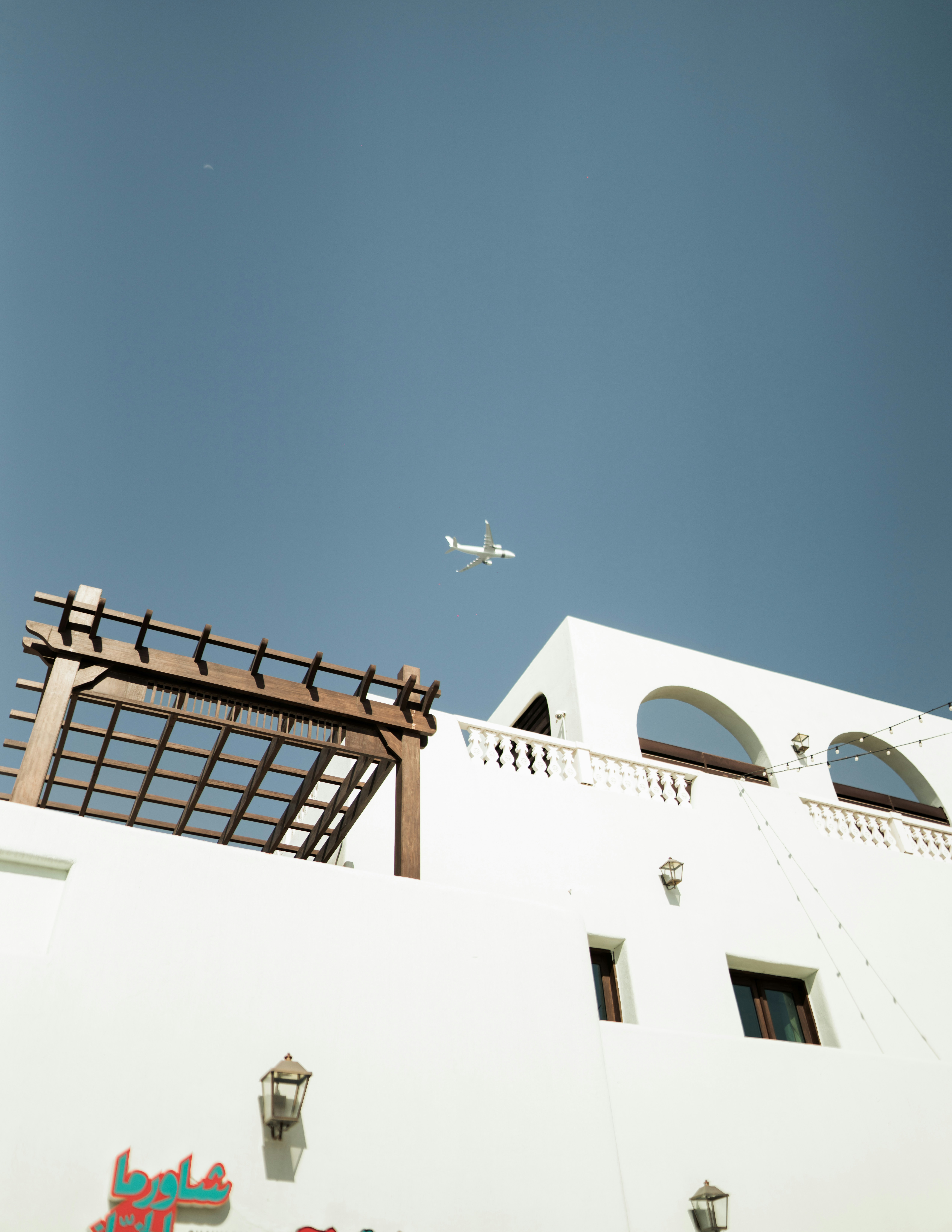 A small airplane flies above a white Mediterranean-style building with a wooden pergola against a clear blue sky.