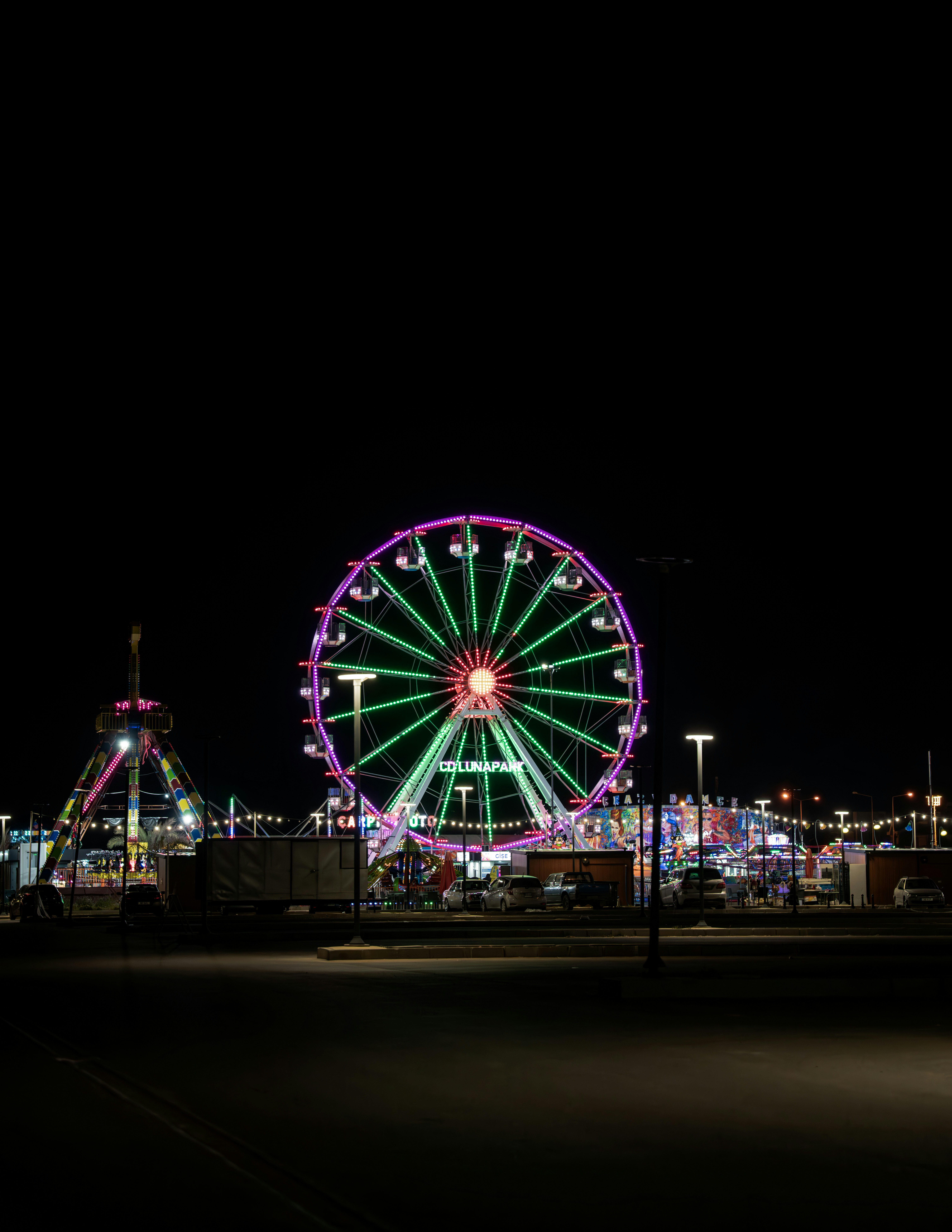 Brightly lit Ferris wheel dominates a night carnival, with colorful spokes and surrounding rides and stalls visible in the dim foreground.