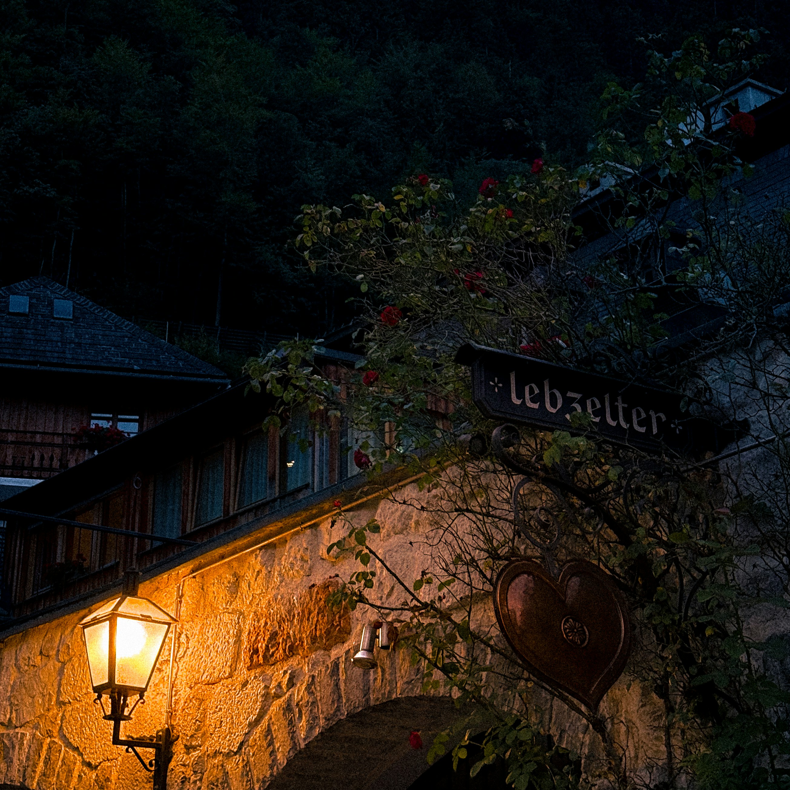 Amber-lit stone archway and streetlamp illuminate a rustic mountain village at dusk; a hanging sign and heart-shaped décor add charm to the scene.