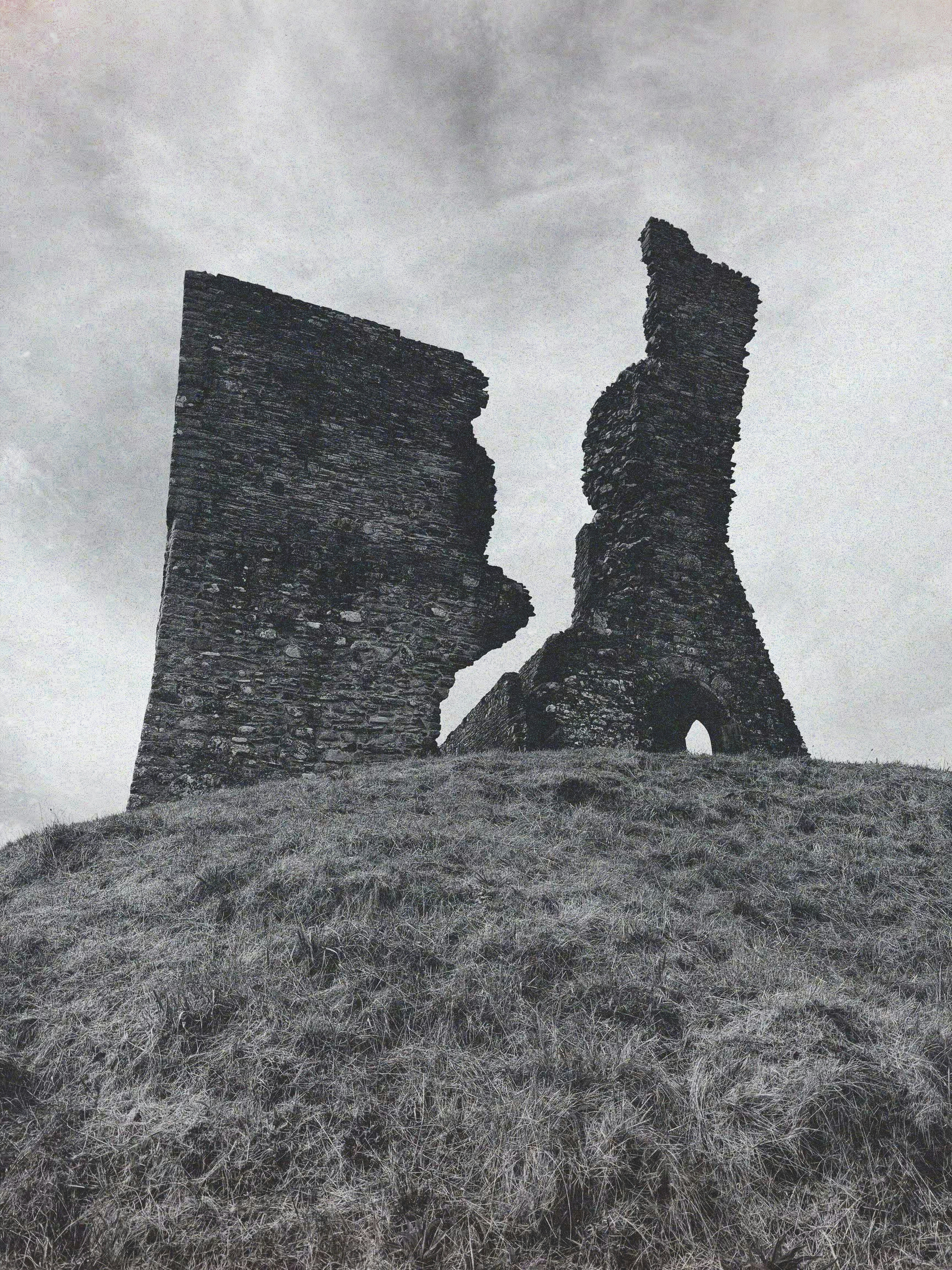 Weathered stone ruins perched on a grassy mound under a dramatic sky, captured in grainy monochrome to emphasize texture and age.