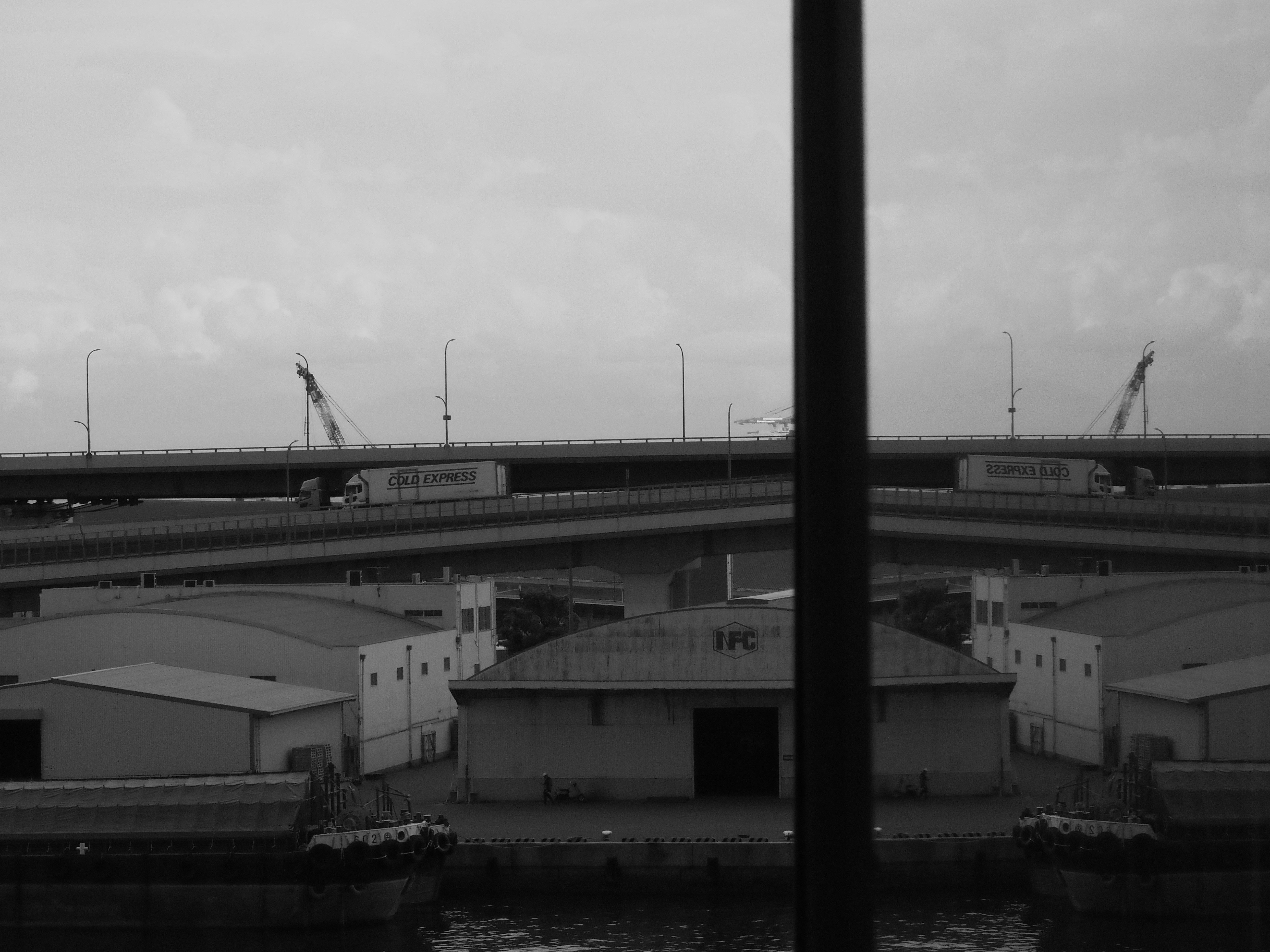 Industrial harbor scene with a central vertical pole dividing a low-contrast landscape of warehouses and a bridge; overcast sky adds softness to the composition.