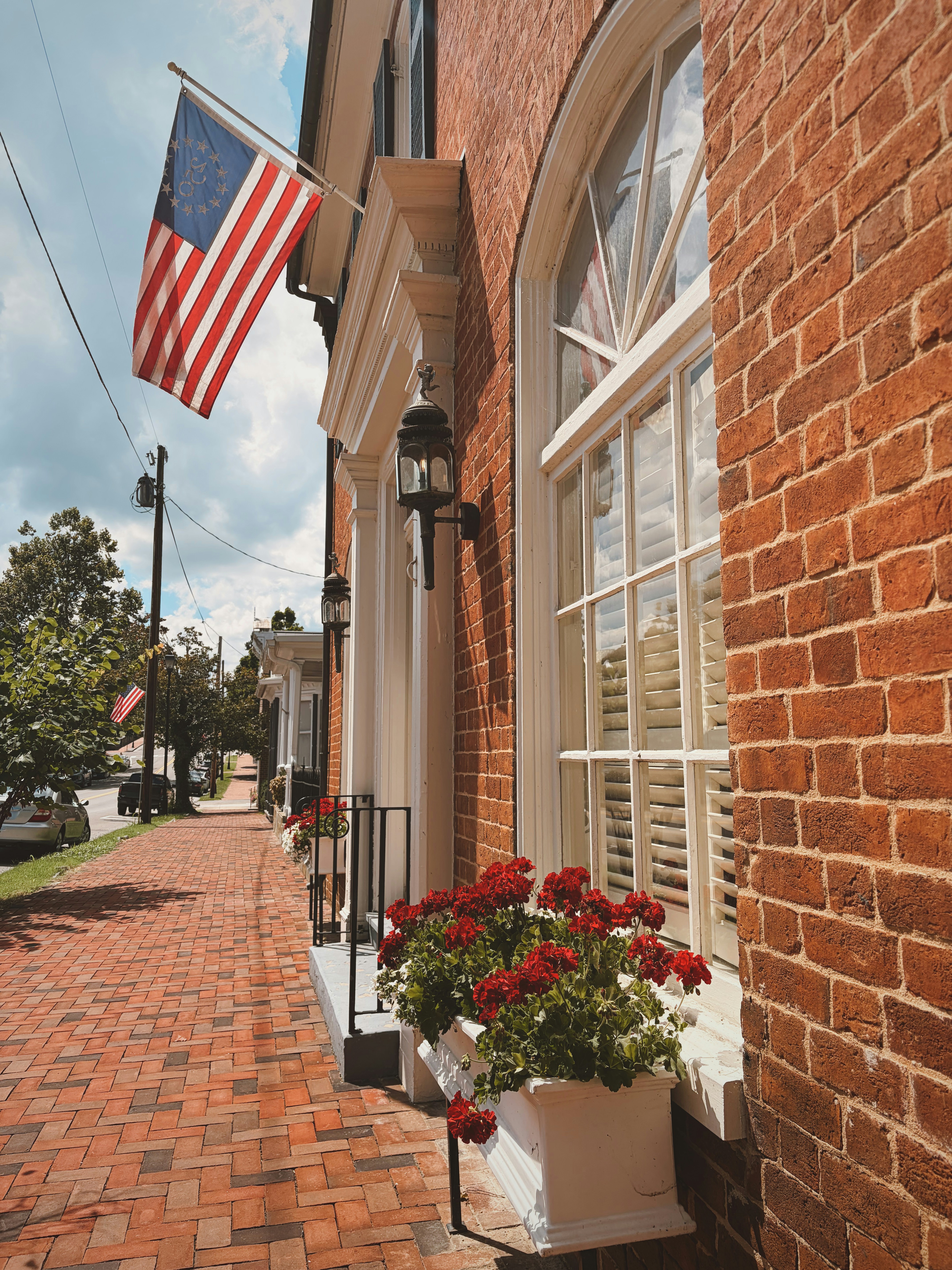 Red brick rowhouse with a white-framed window and hanging lanterns, a planter of red flowers, and an American flag along a brick sidewalk.