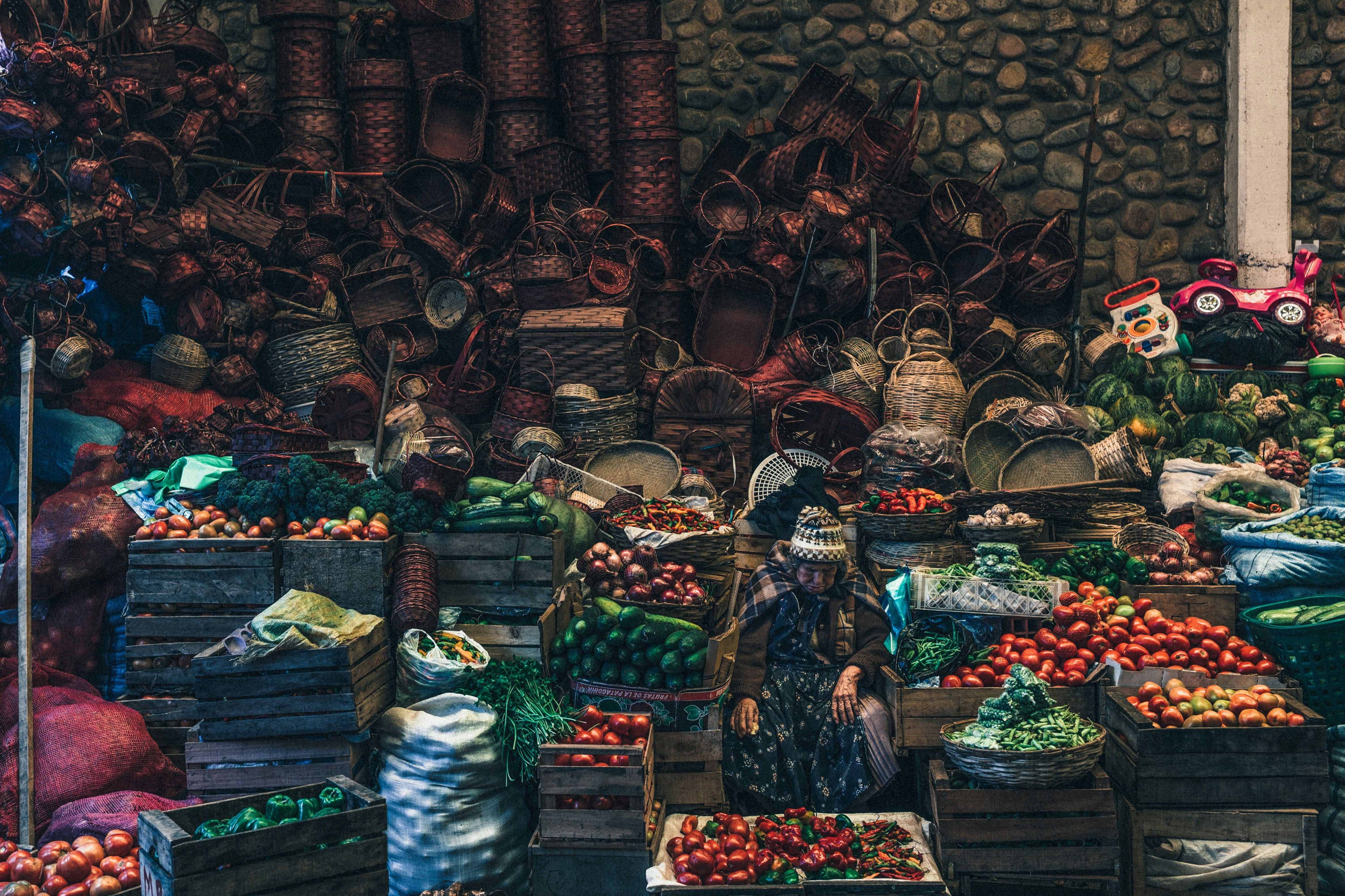 Colorful market stalls with crates of vegetables and a vendor amid woven baskets in a crowded alley.