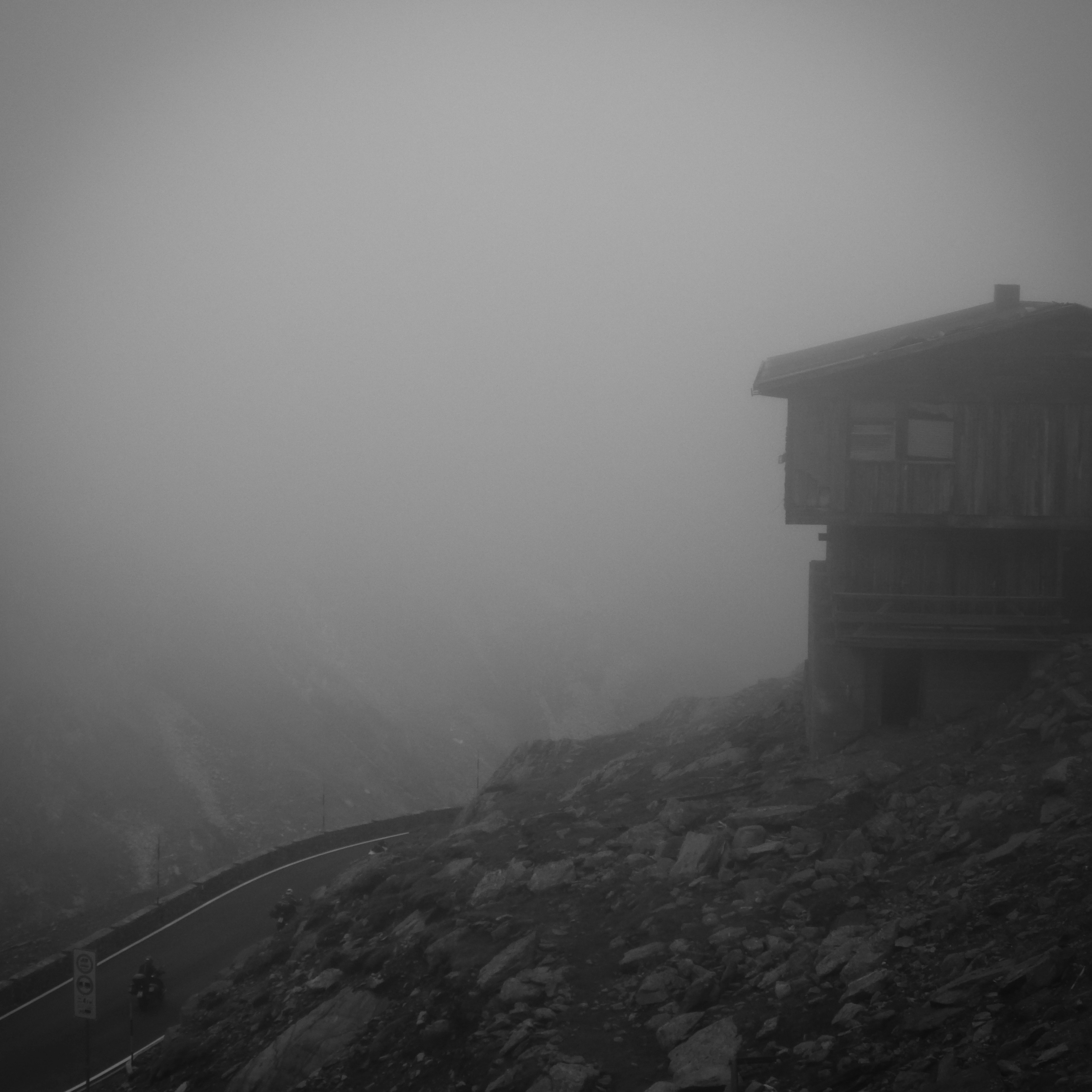 A solitary wooden cabin sits on a rocky slope beside a winding road, shrouded in dense fog that obscures the distant landscape.