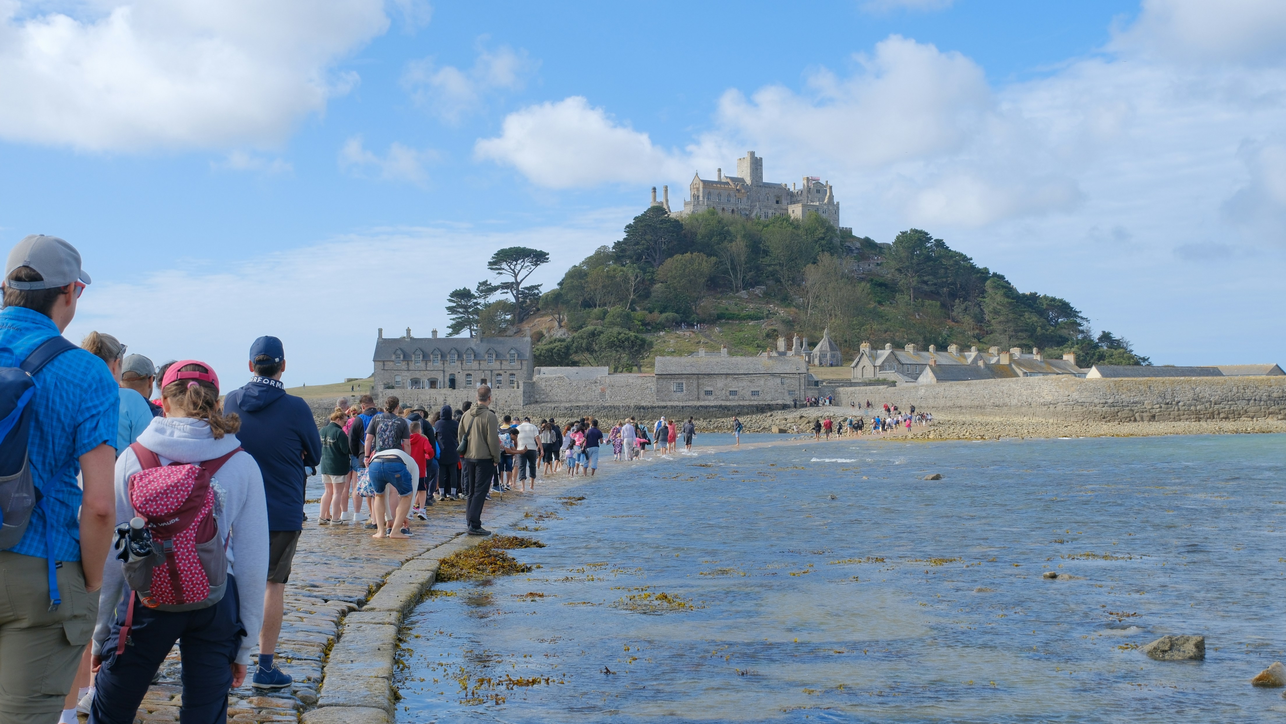 Crowds Walk a Sea‑Front Path Toward a Hilltop Fortress