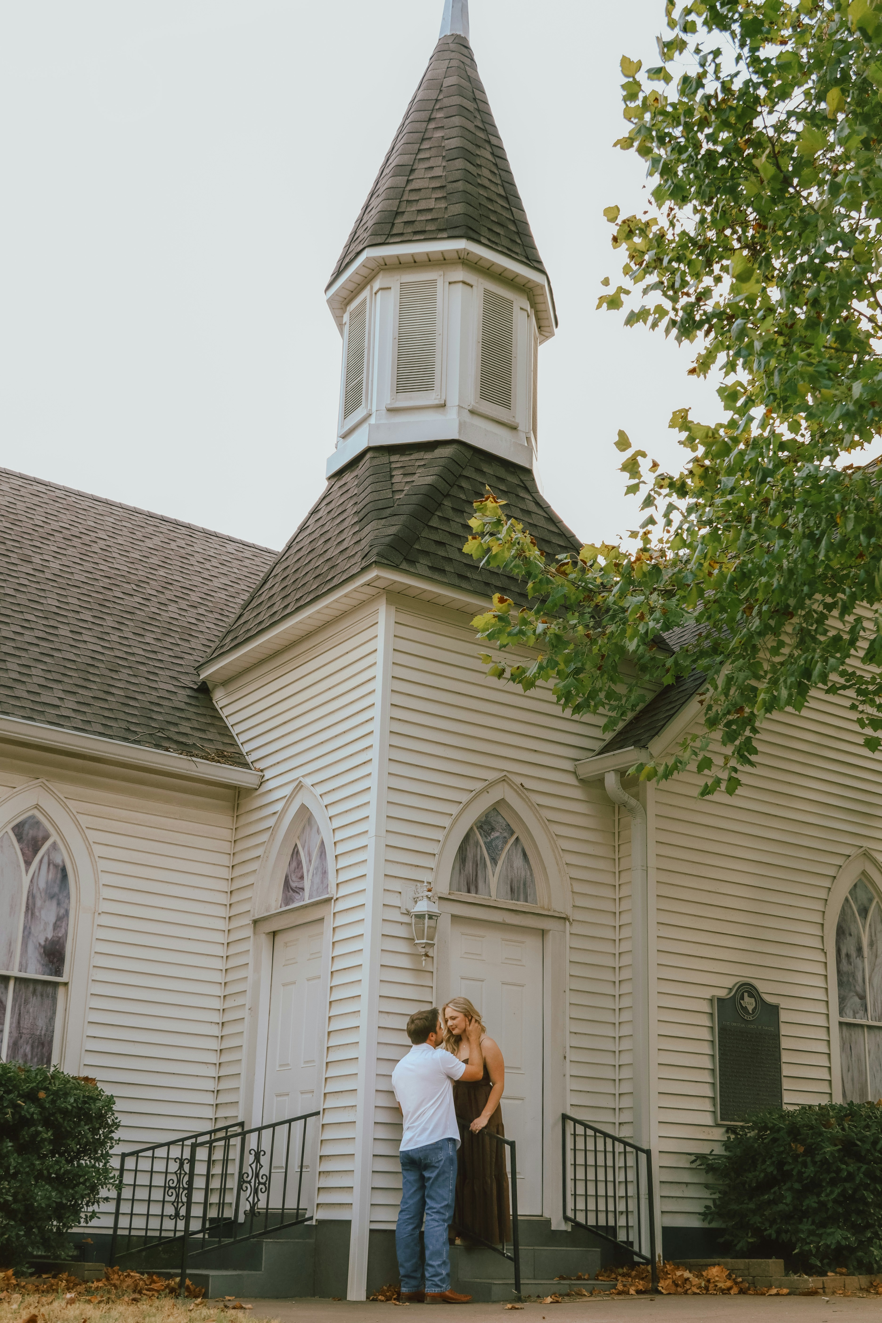 A couple shares a tender moment at the base of a white church with a tall steeple, framed by green trees and autumn leaves along the steps.