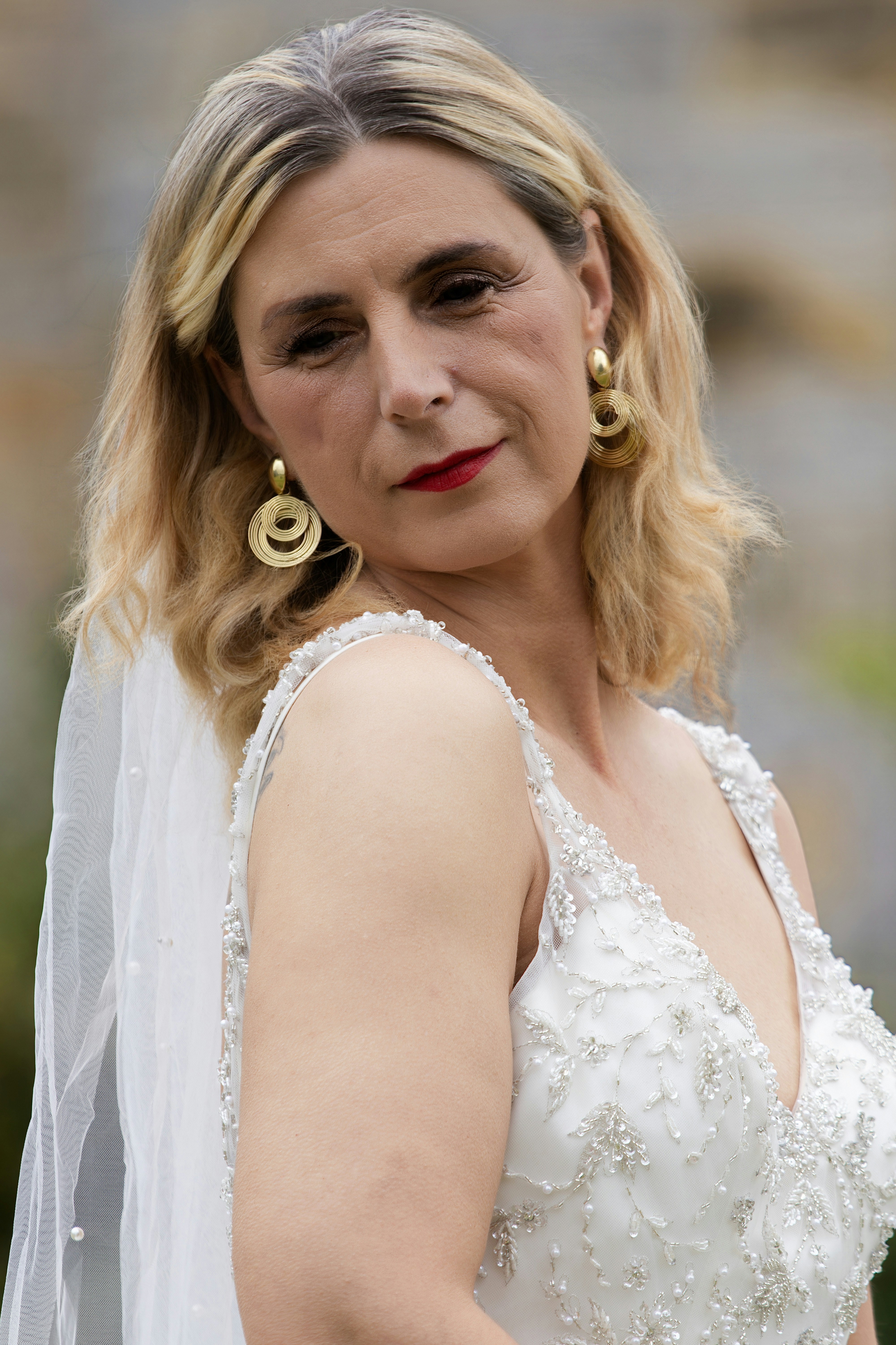 Elegant close-up of a woman in a lace bridal gown, her face slightly angled as she gazes downward with soft makeup and gold earrings. The shallow depth of field keeps the subject sharp against a muted background.