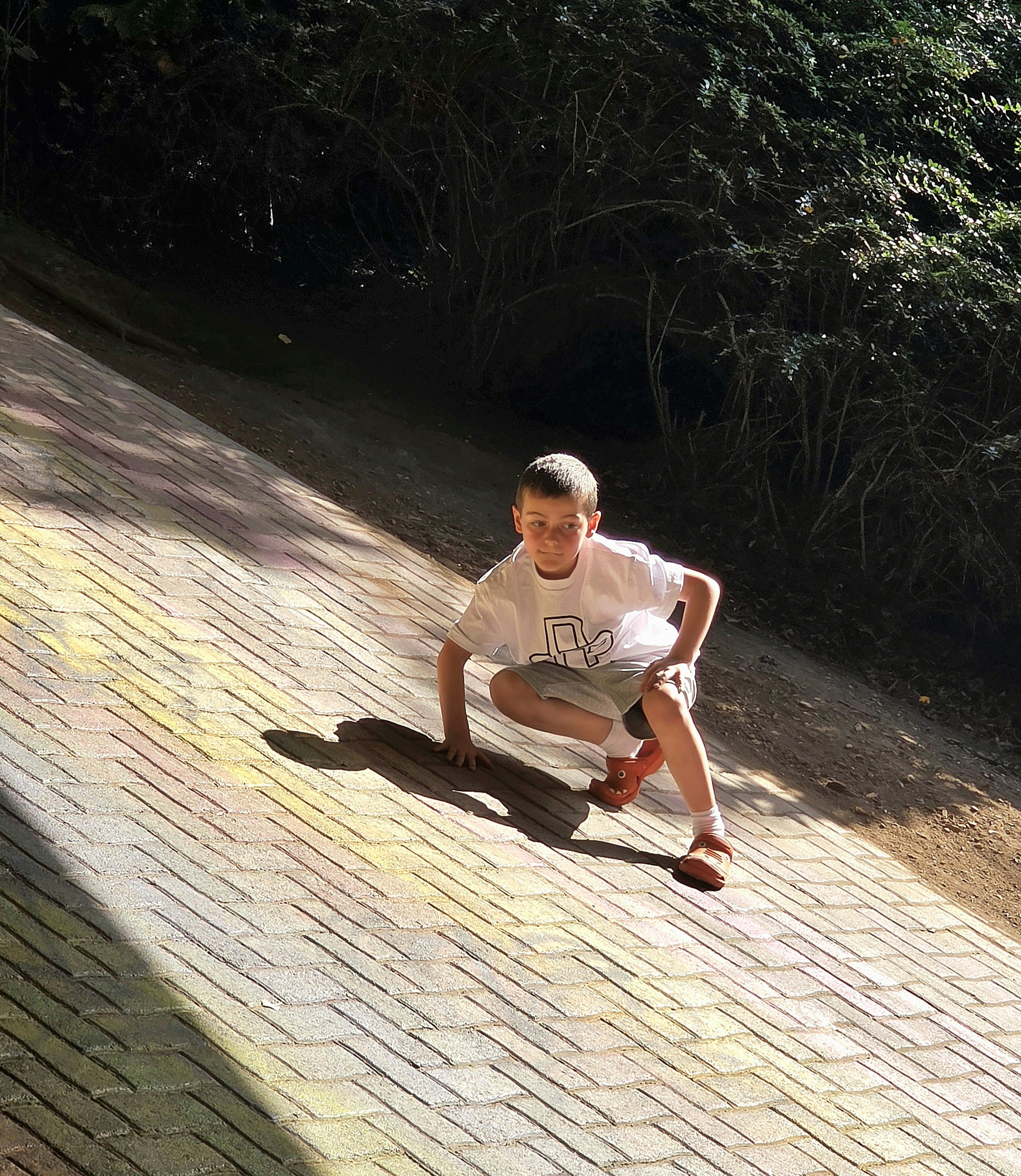 Boy crouches on sunlit brick path, shaded hillside framing a quiet moment