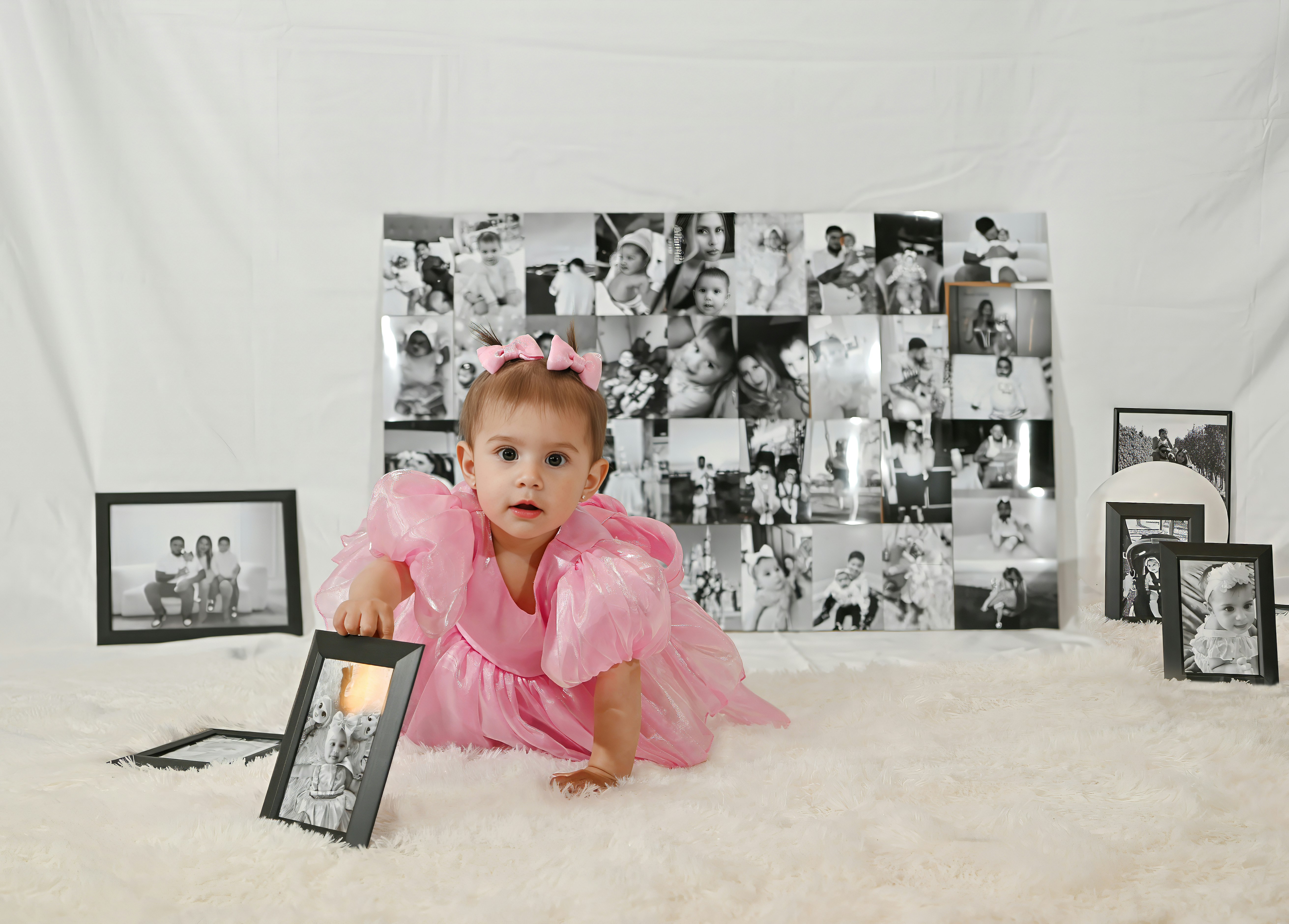 A baby in a pink tutu sits on a soft white surface, framed by black-and-white photos arranged on a backdrop and scattered frames around her.