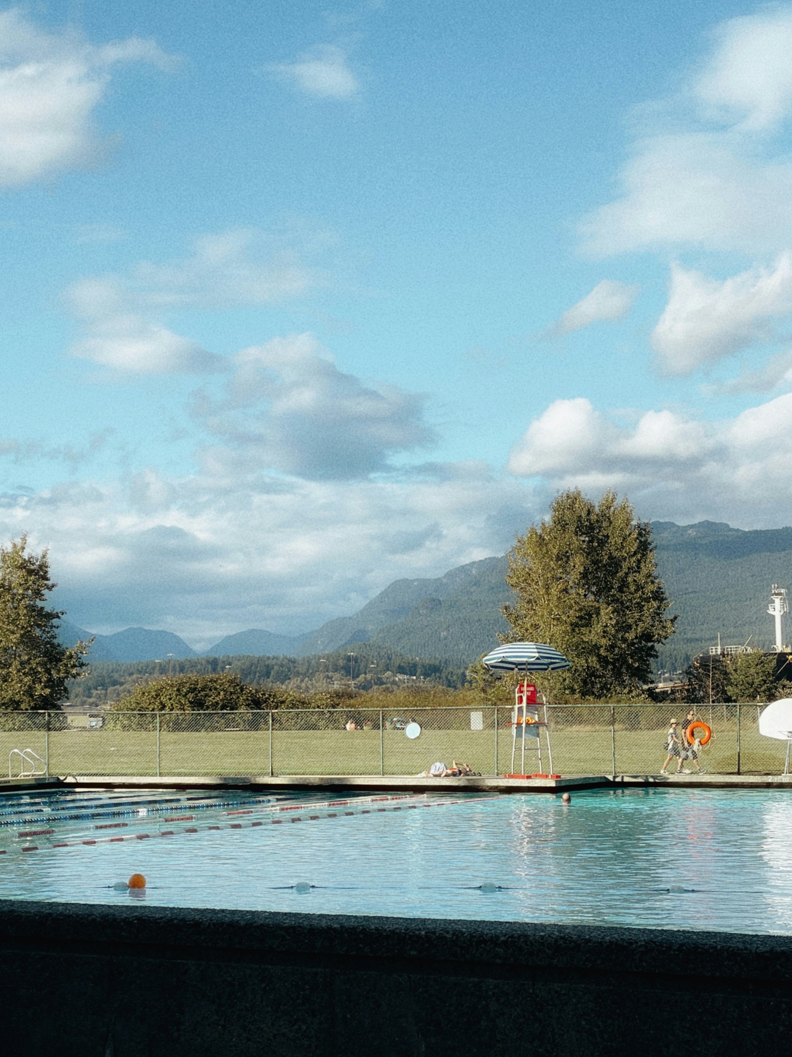 A tranquil outdoor pool scene with blue sky, distant mountains, and a lifeguard chair by the pool edge. The setting captures leisure and calm in a wide landscape.
