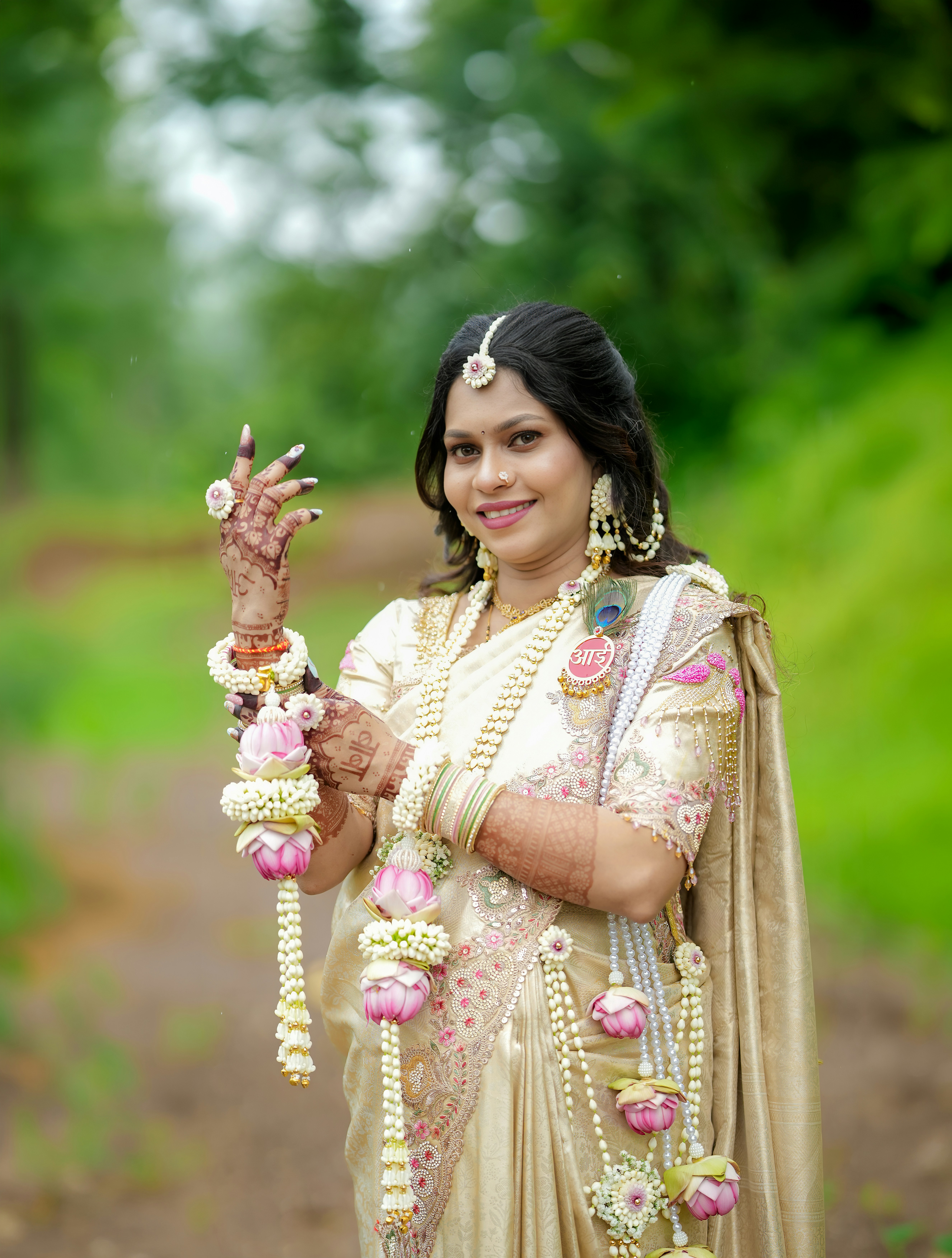 Richly dressed bride-adorned with floral garlands poses outdoors, focusing on her ornate jewelry and henna-adorned hands; the image emphasizes ceremonial detail and natural backdrop. (Logo: none, Illustration/Painting/Cartoon: none).