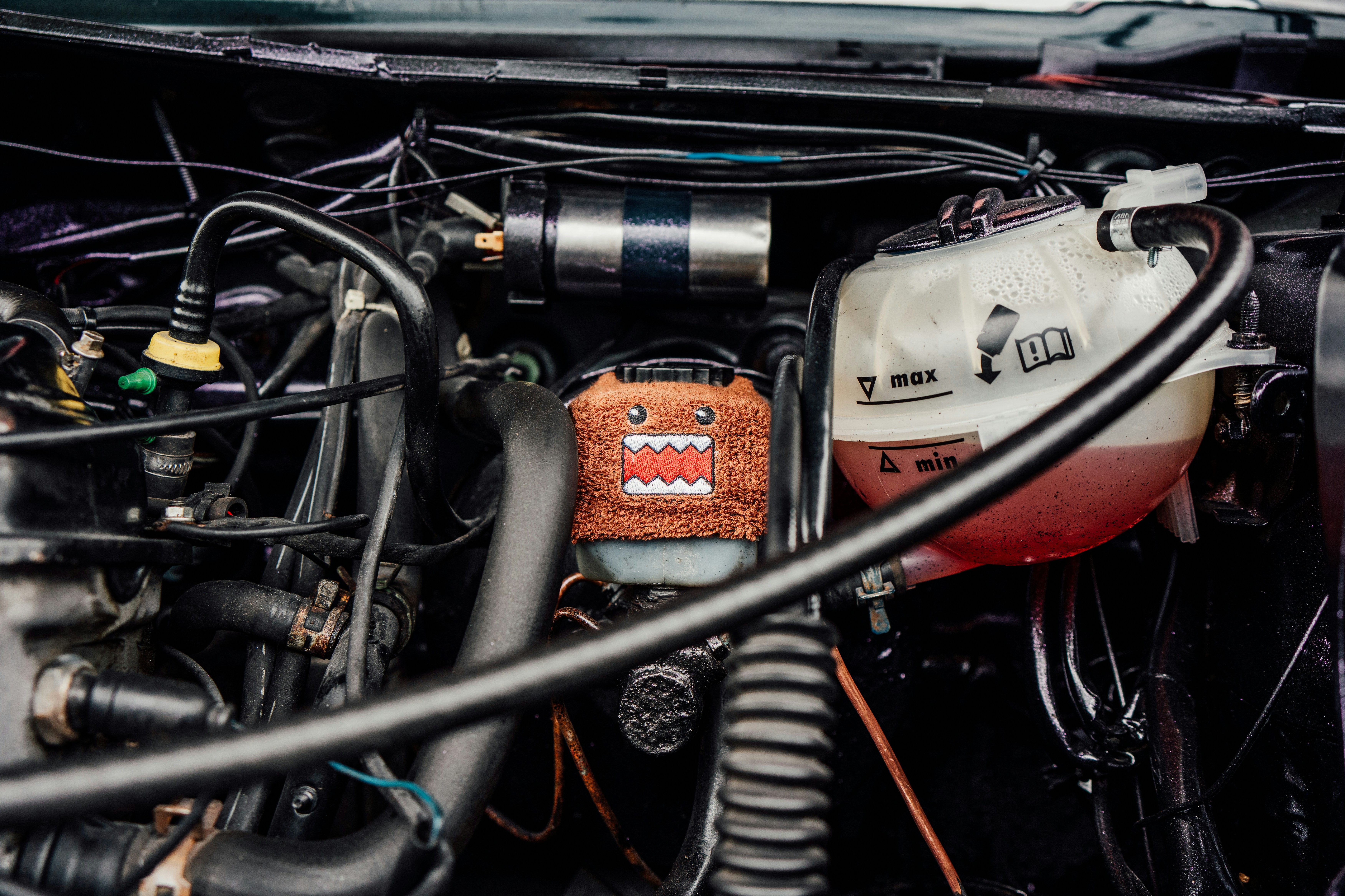 Close-up of a car engine bay featuring a red-orange ignition coil and surrounding hoses and reservoirs; industrial look with exposed components and wiring. This is a photo of machinery, not a logo or illustration.