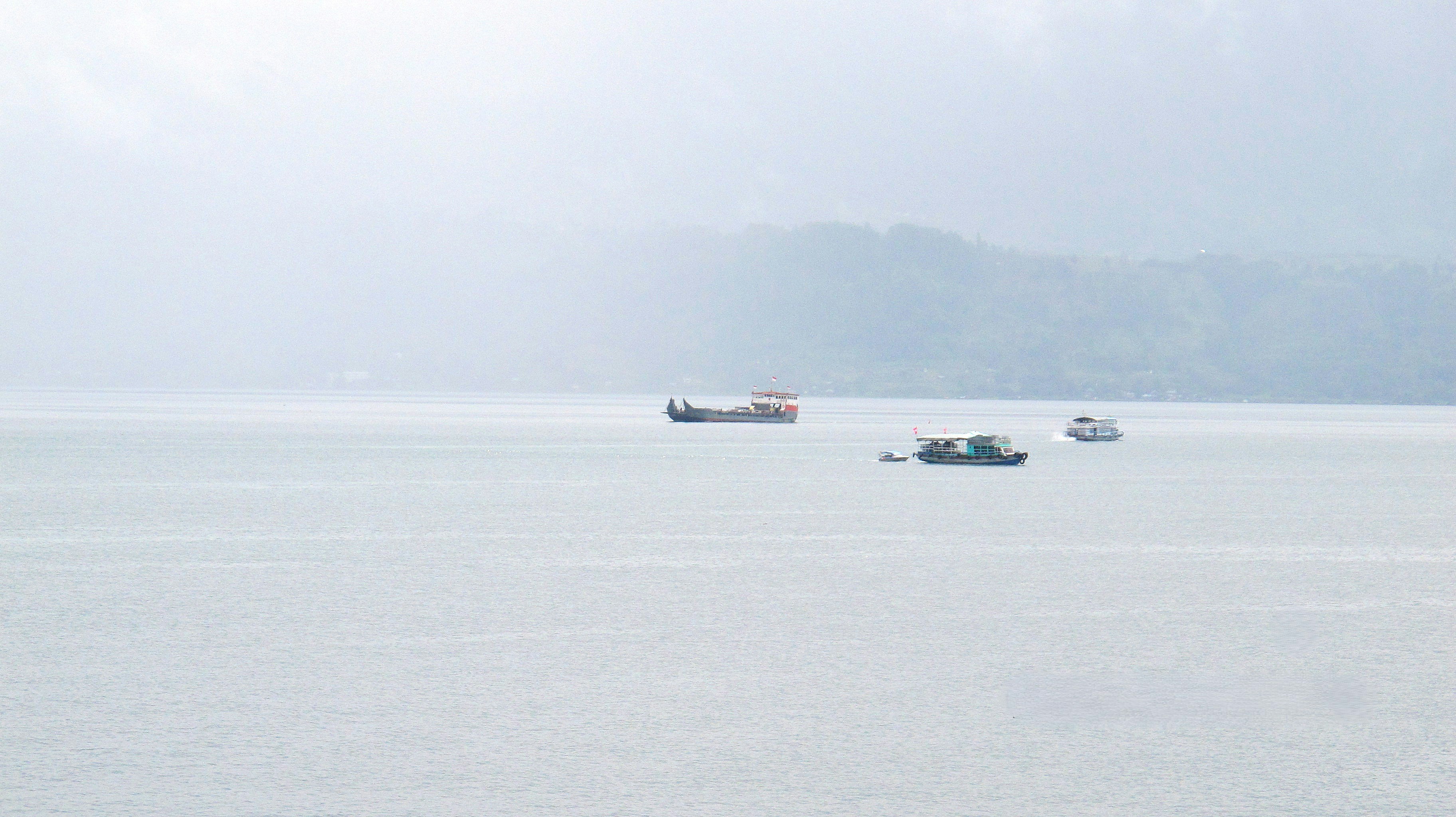 Softly lit harbor scene with a few distant boats on pale gray water under a misty sky. Abstract calm seascape emphasizing the horizon and distant hills.