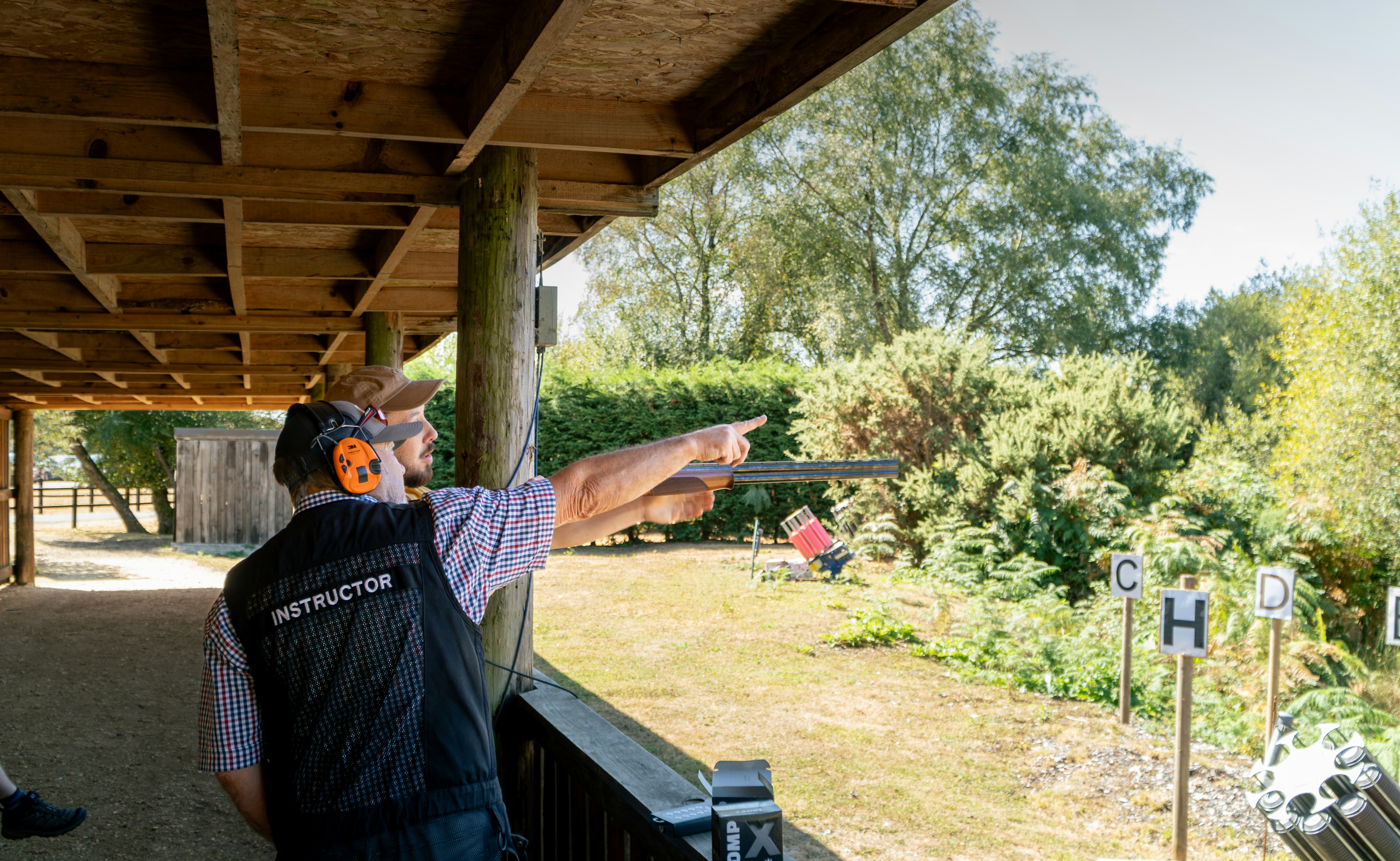 Clay pigeon shooting in the New Forest, England — instructor-led session on a sunny day amid greenery and wooden shelters.