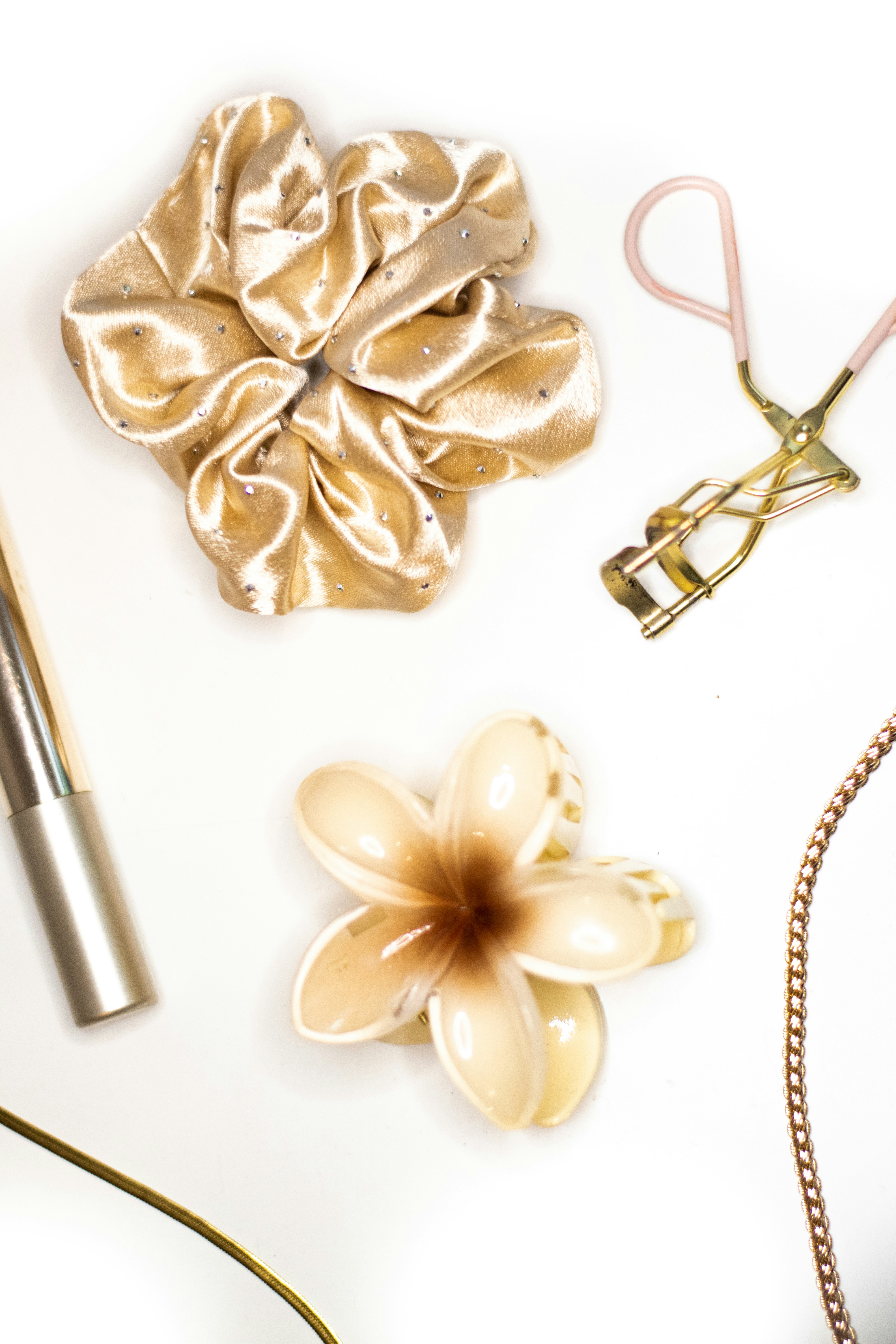 Bright flatlay featuring gold-toned scrunchie, flower-shaped accessory, makeup pencil, and jewelry on a white background. A minimal beauty desk composition with soft shadows and warm metallic tones.