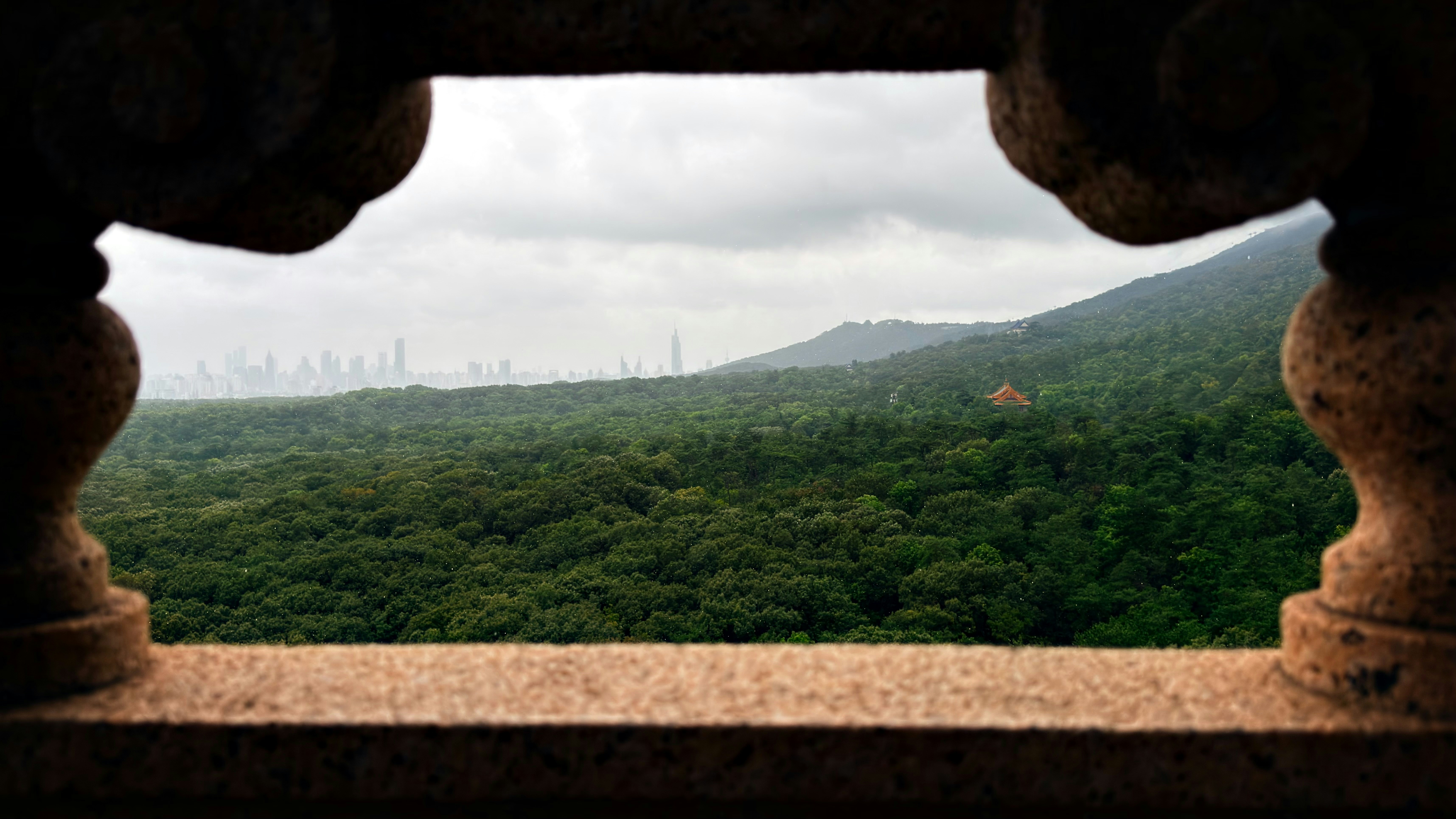 Lush forest stretches to distant hills framed by a stone window, with a faint skyline beyond the trees. A calm, panoramic vista emphasizing depth and natural texture.