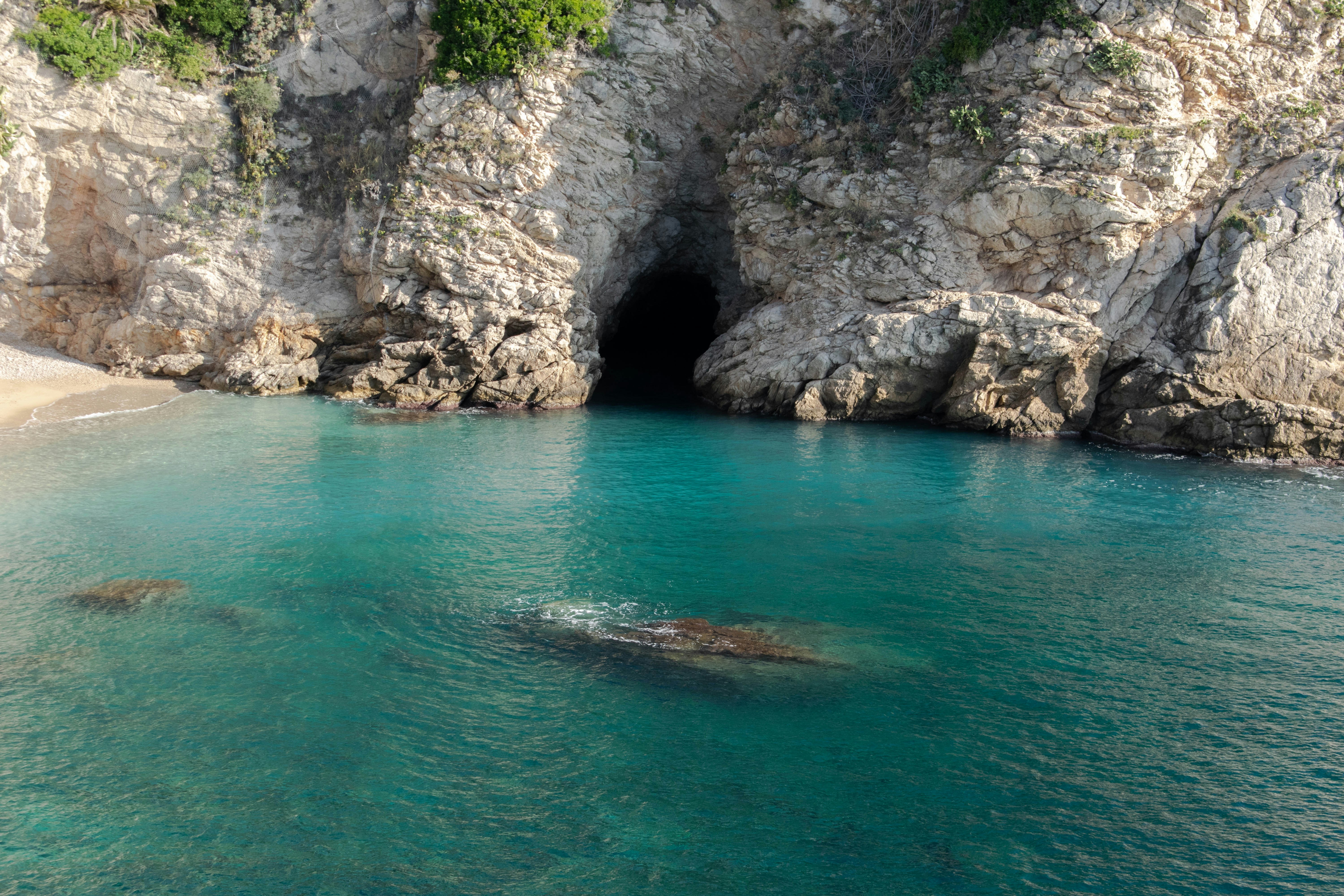 Turquoise sea laps a rocky shoreline as a dark sea cave opens from the cliff, with clear water and a small sandy beach on the left.