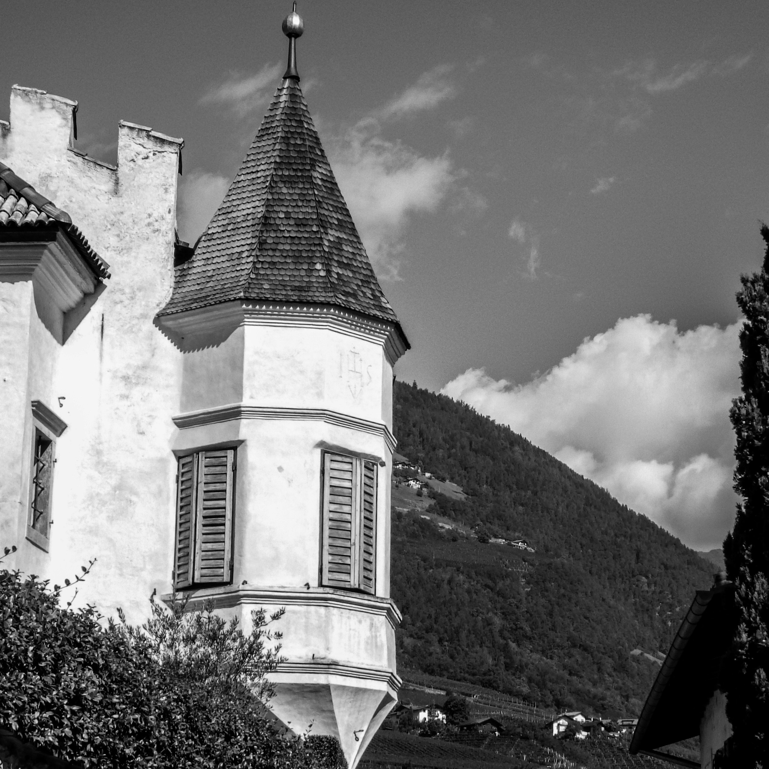 A grayscale photograph of a classical castle turret with shuttered windows, perched against a mountainous landscape and cloudy sky. The scene emphasizes the architectural detail and surrounding natural backdrop.
