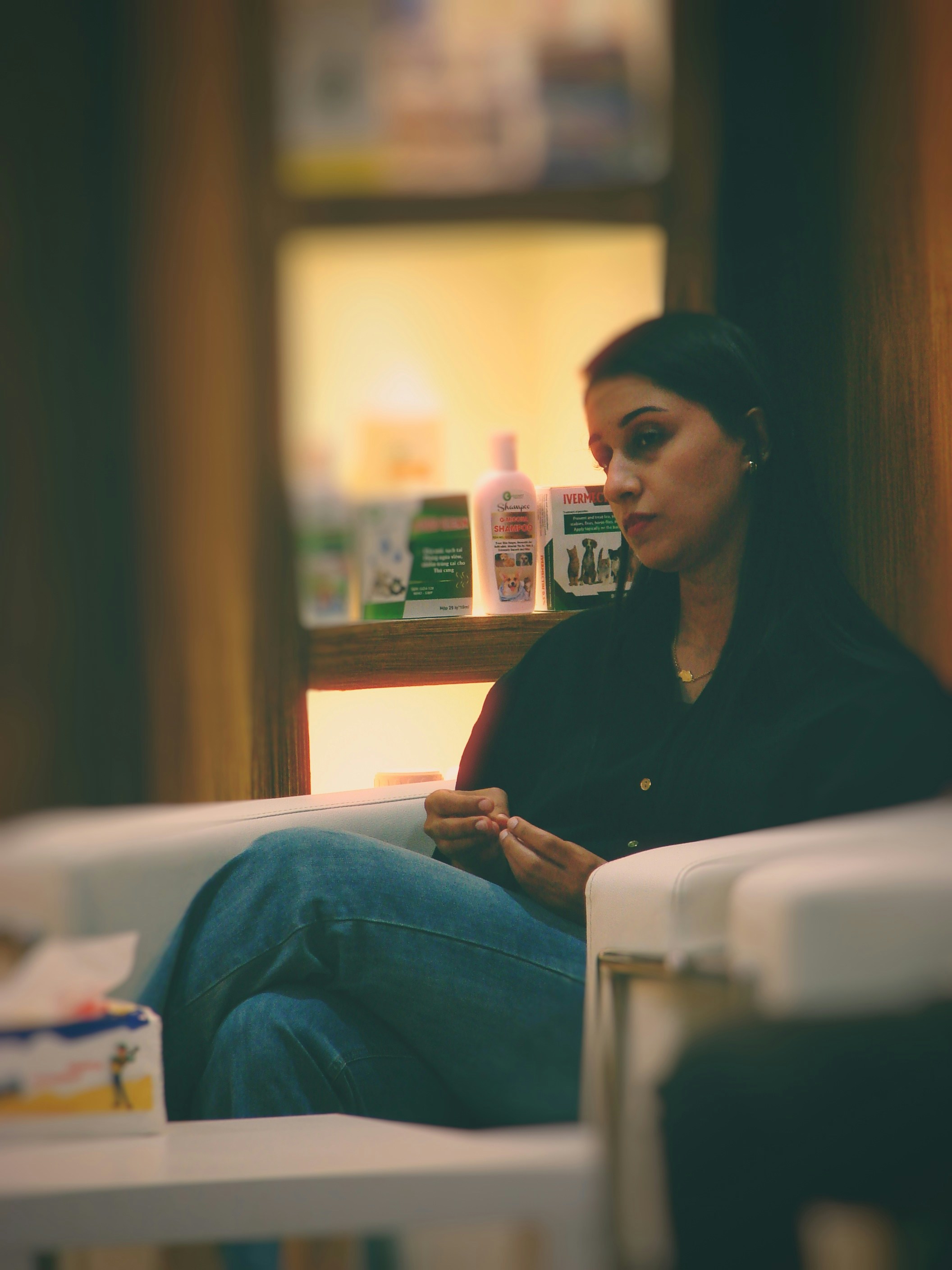 A woman sits in a softly lit, cozy lounge with a relaxed posture, the scene focused on her calm expression and hands resting in her lap.